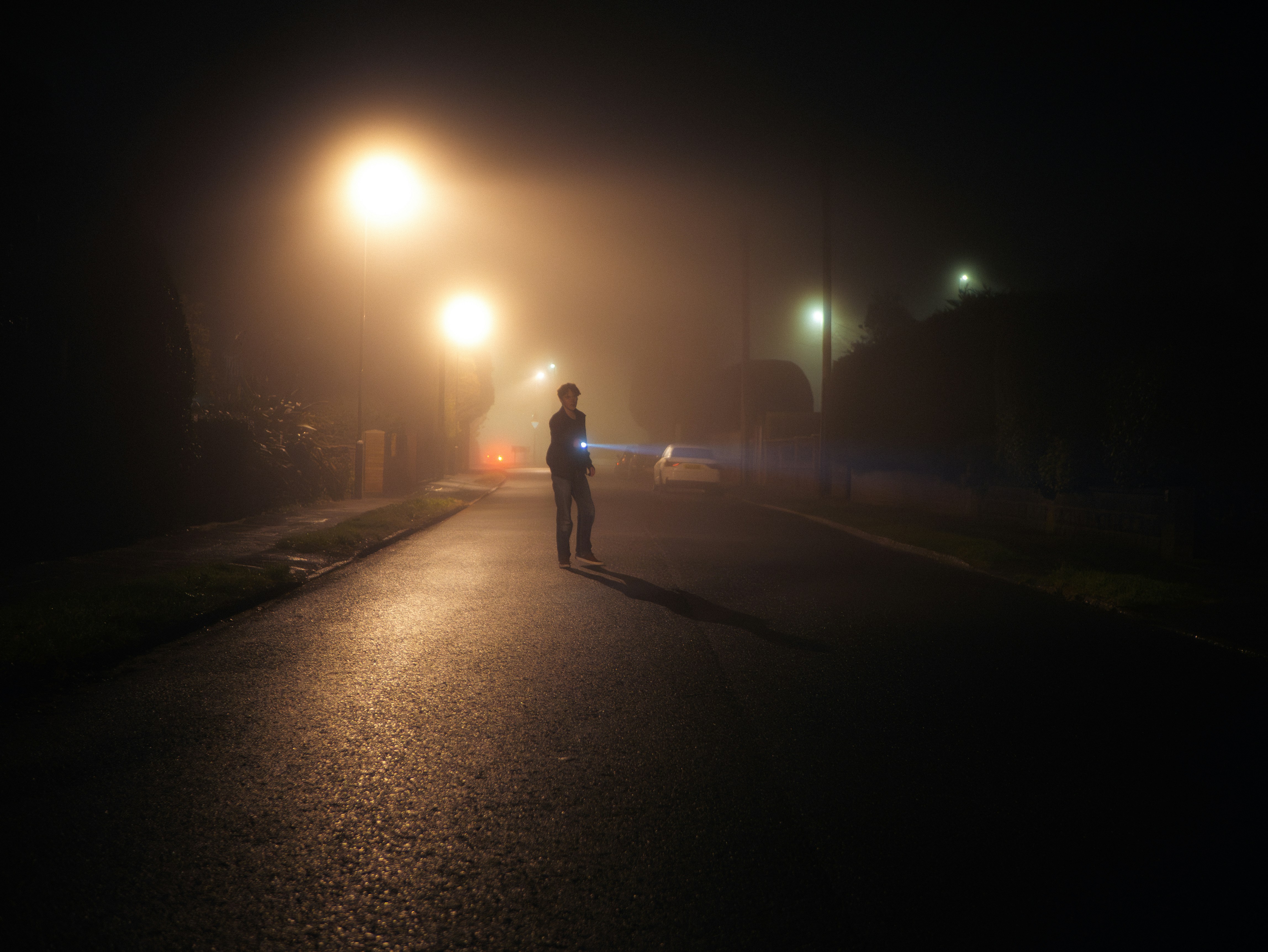 Solitary figure walks on a foggy street illuminated by warm streetlights, casting long shadows on wet pavement.