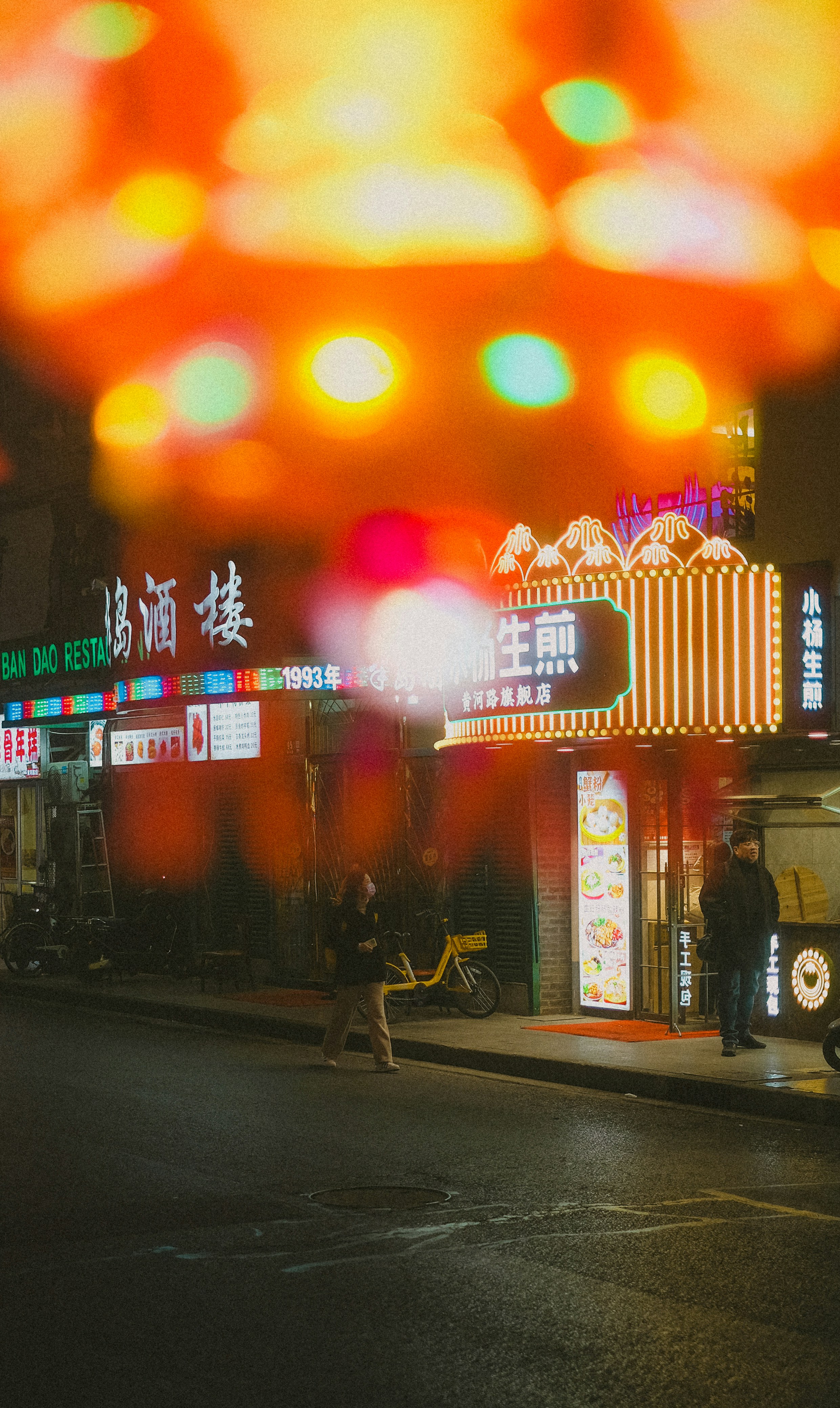 Night street scene with neon signs and blurred lantern lights above a row of storefronts. Pedestrians stand near glowing entrances, hinting at a late-night market.