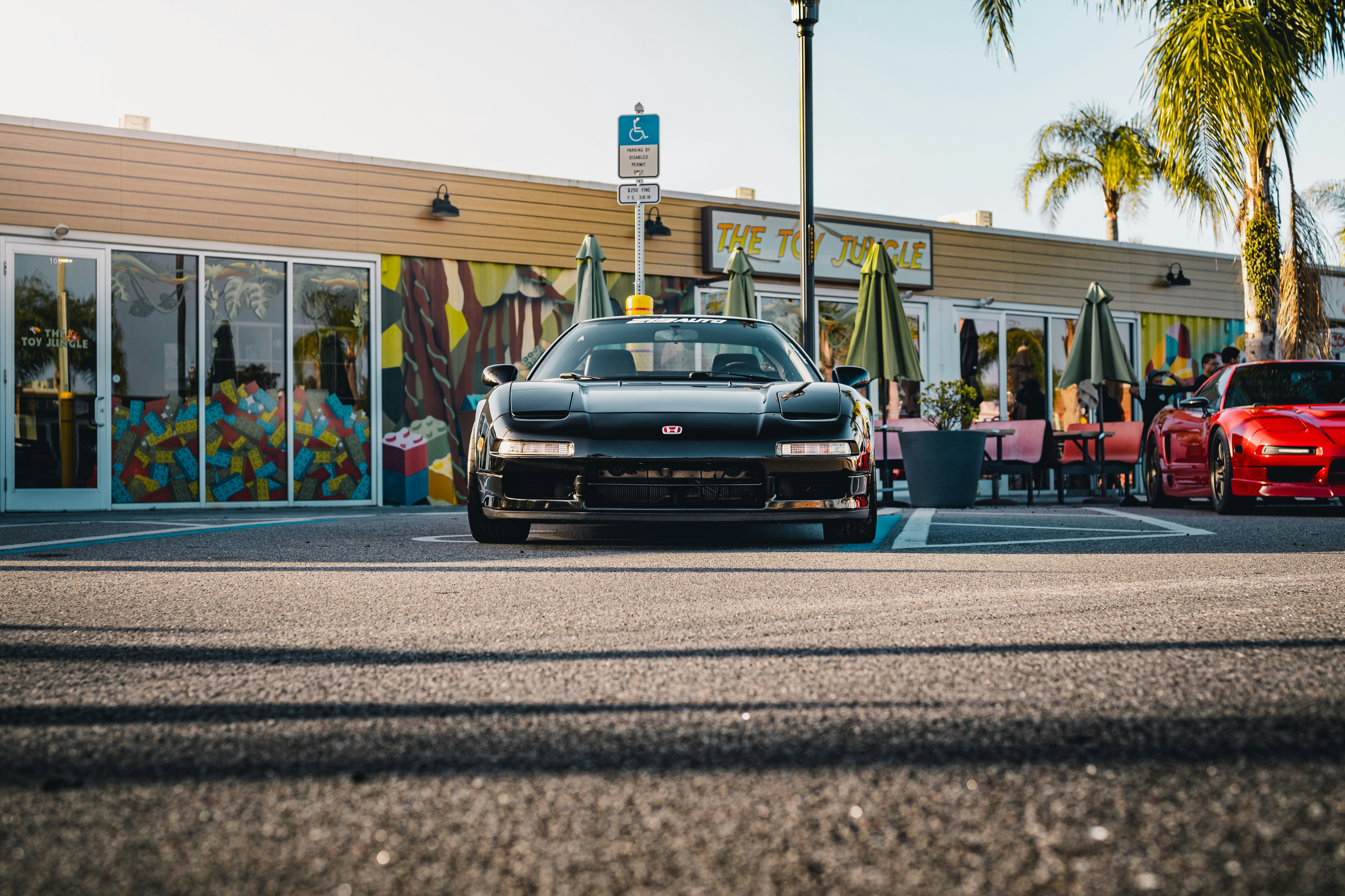 Black sports car front and center with a vibrant storefront backdrop and a red car nearby. Palm trees and long shadows enhance the scene.