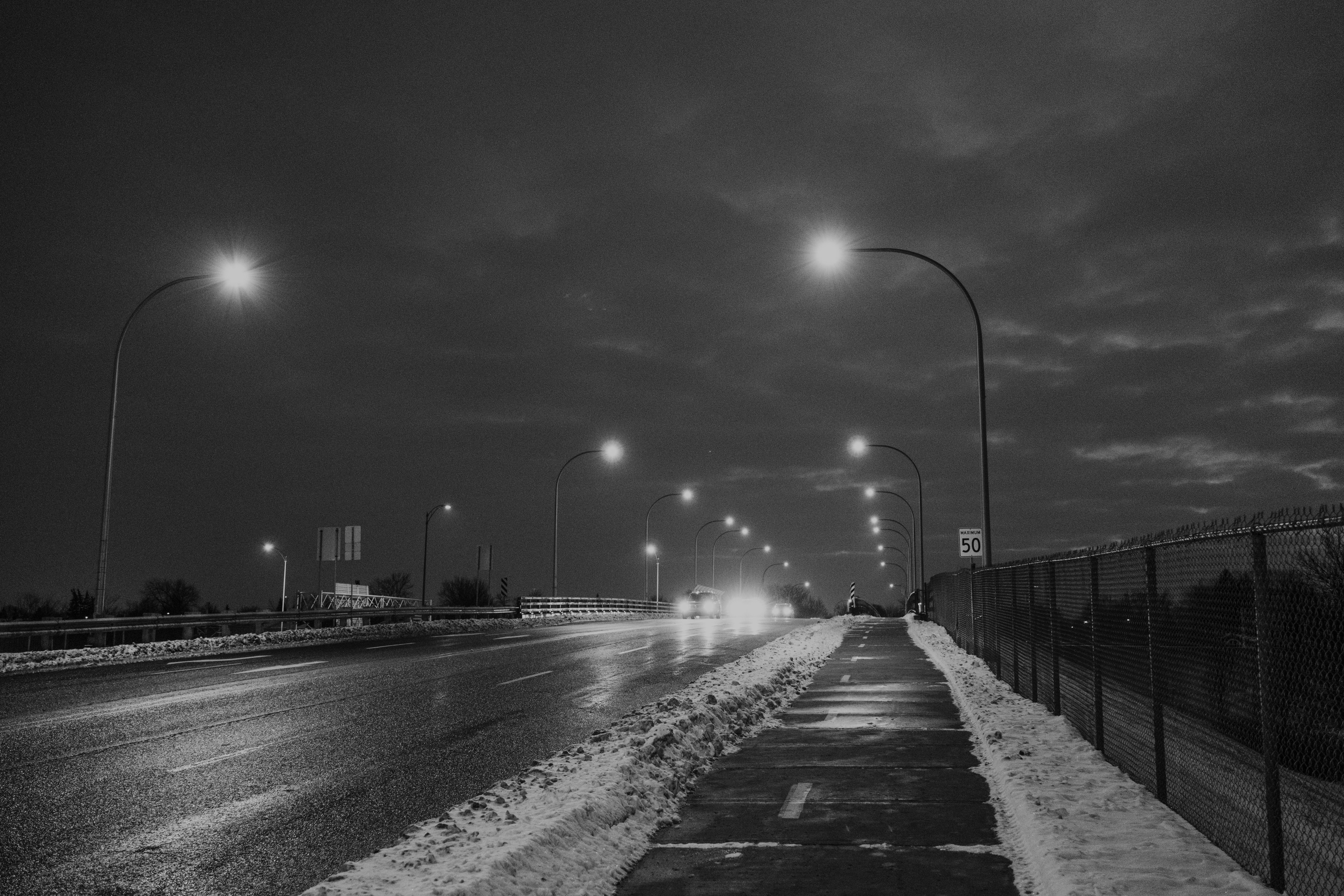Black and white urban street at night with glowing streetlights lining a snowy sidewalk, casting reflections on the wet road.