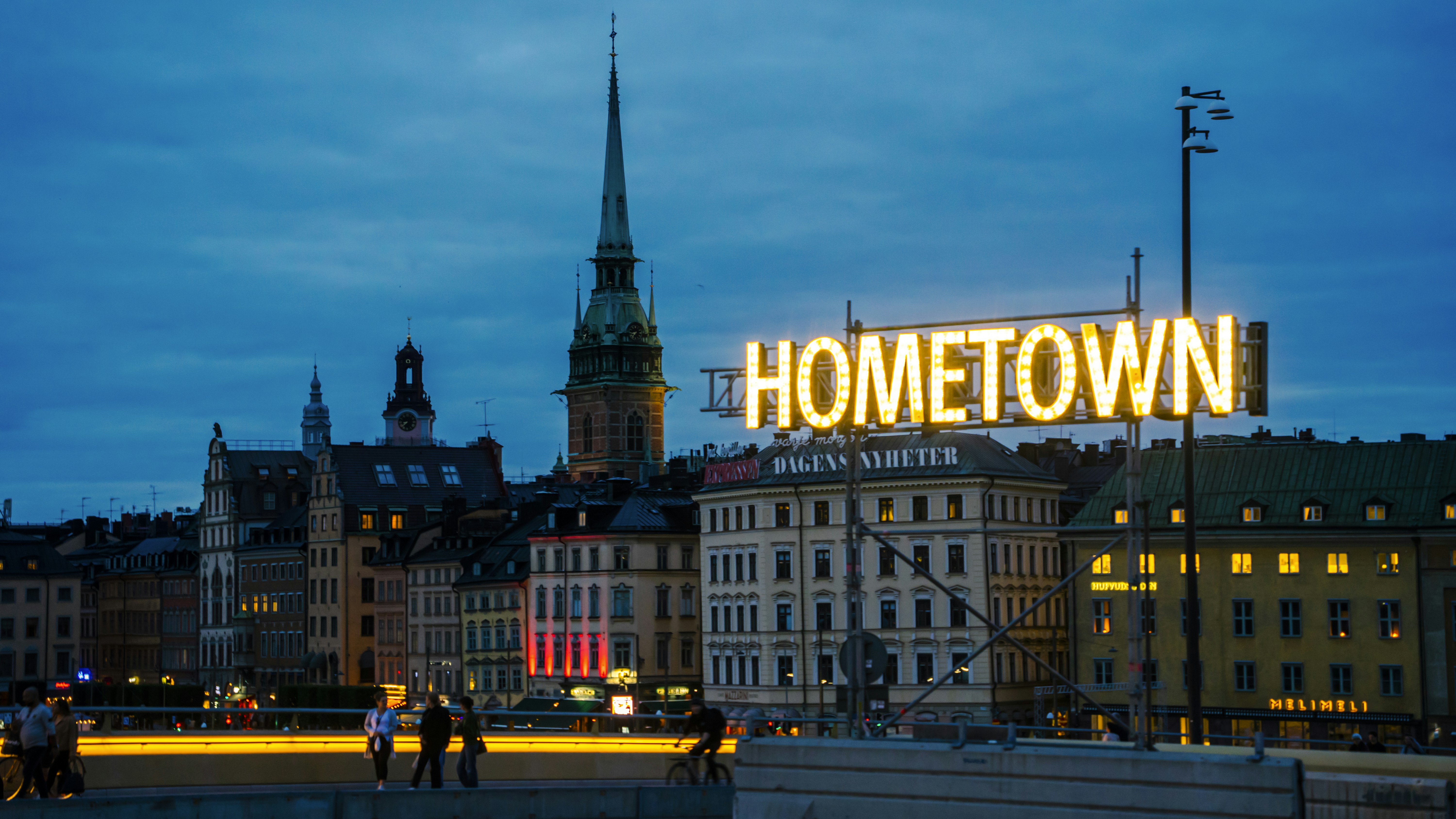 Illuminated sign reading 'HOMETOWN' against a backdrop of historic buildings and a church spire at dusk.