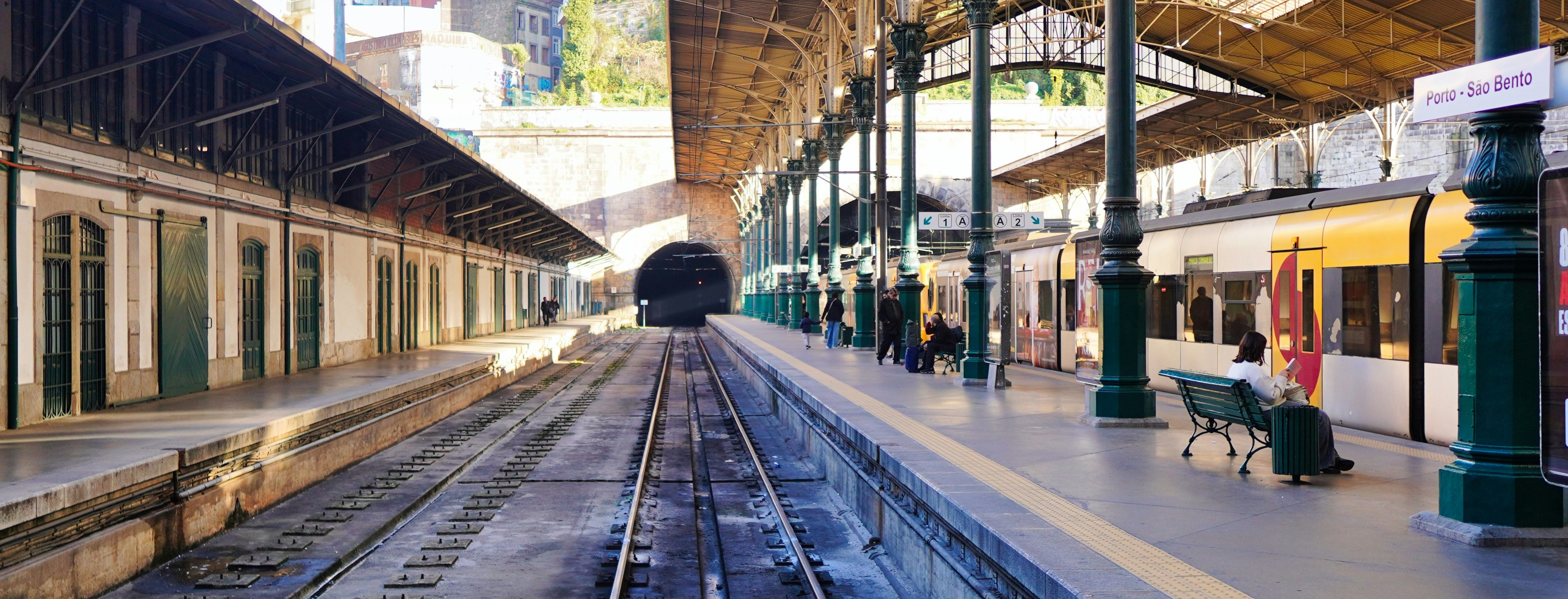 Train tracks leading into a sunlit tunnel at São Bento station, with passengers waiting on the platform.