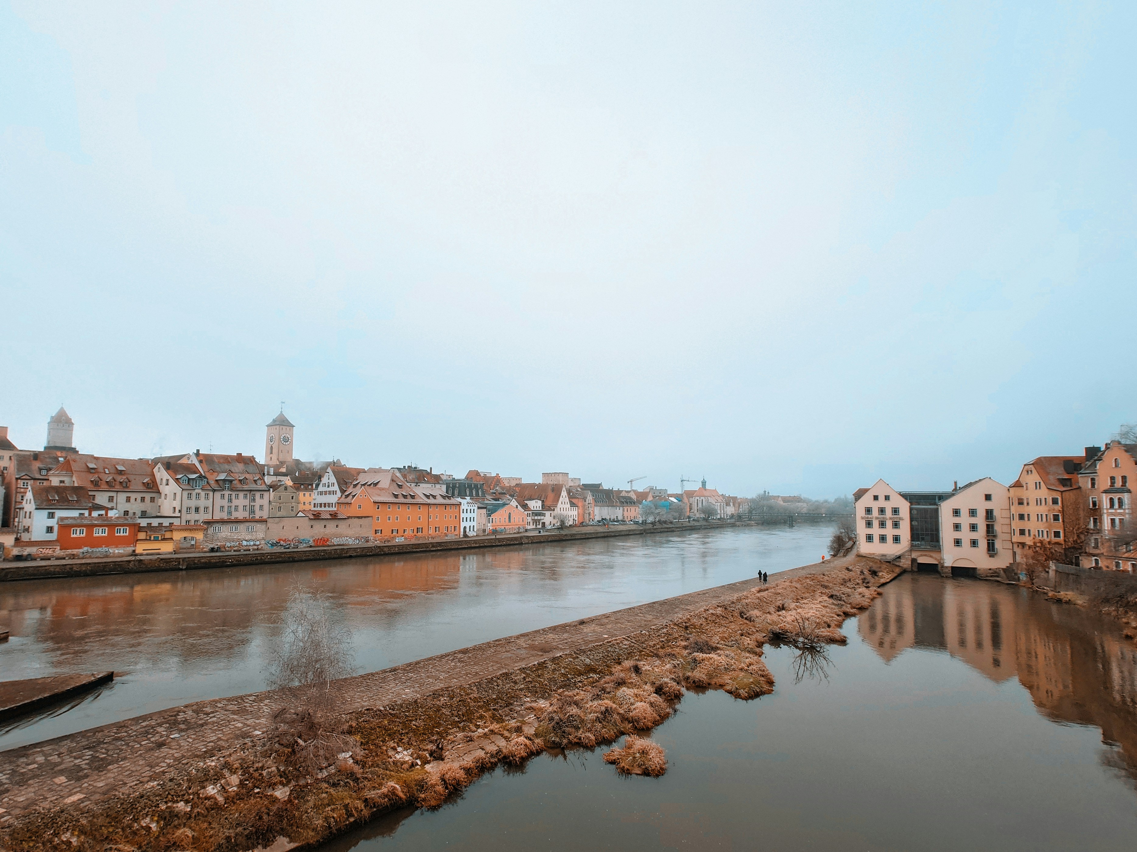 Calm riverfront of a European town with red-tiled roofs and stone quays mirrors itself in the water. A tranquil morning mood emphasizes historic architecture and quiet waterfront life.