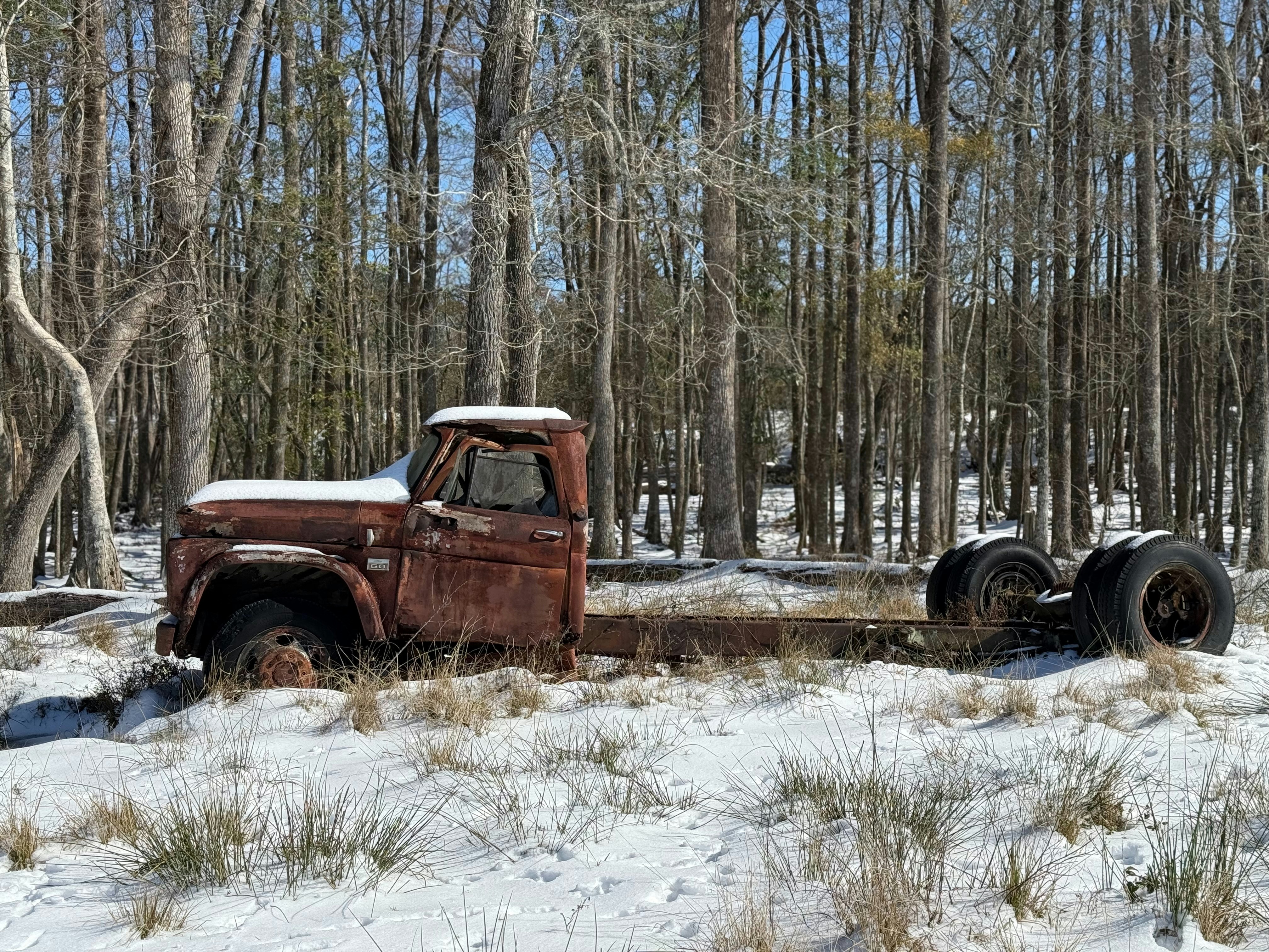 A rusted spray truck partially buried in snow within a quiet forest.