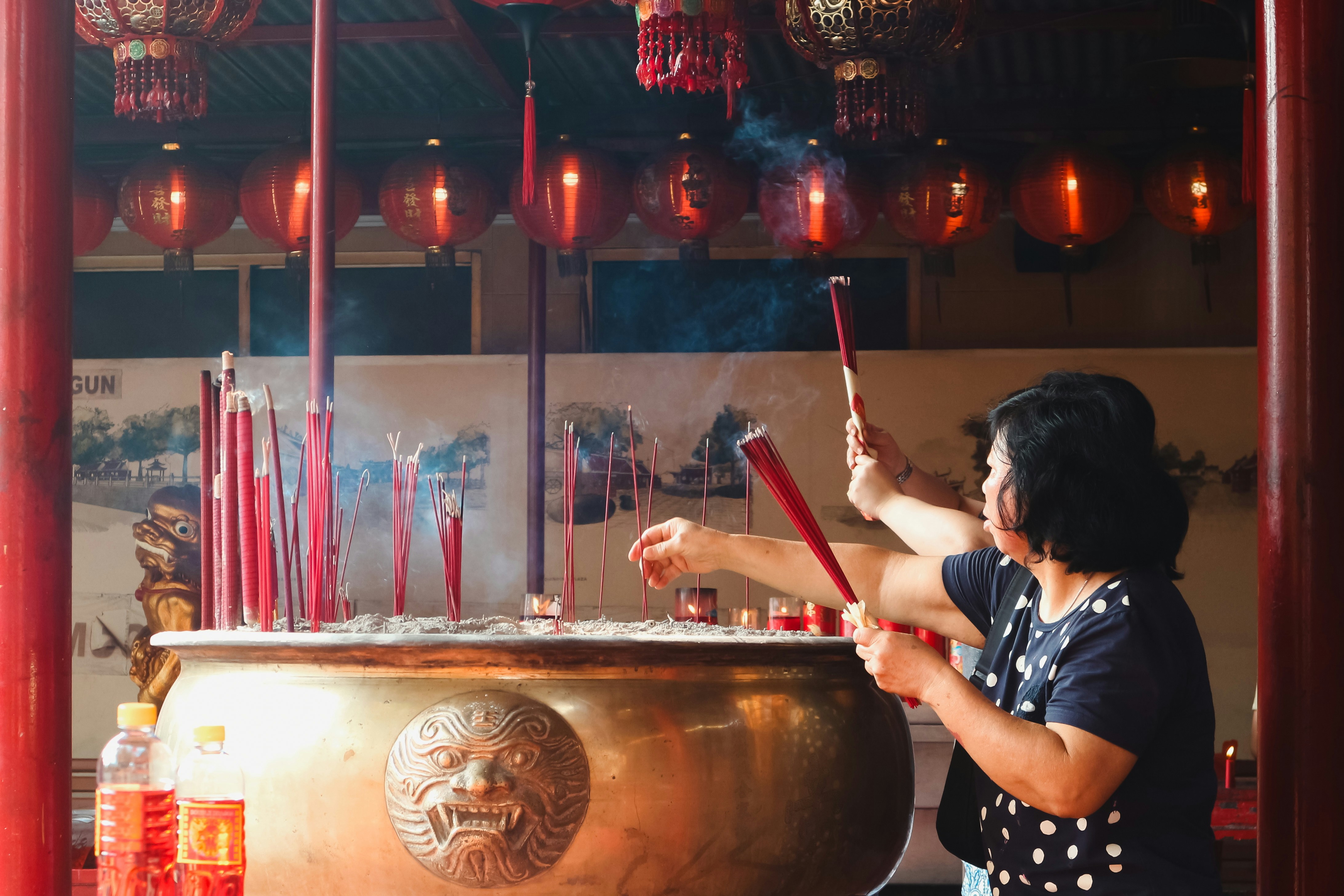 Person placing incense sticks in a large ornate brass urn surrounded by red lanterns and smoke.