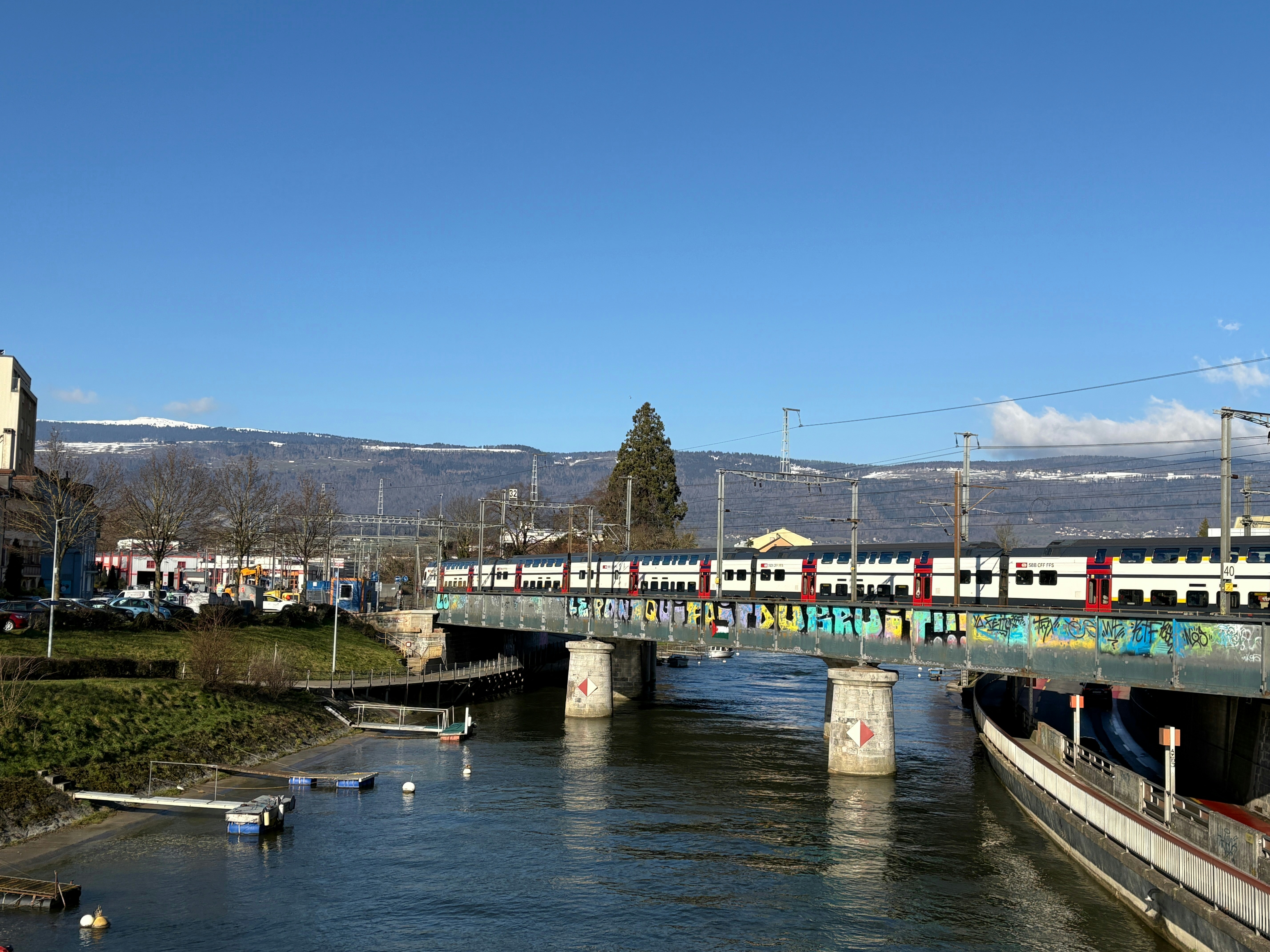 Passenger train crossing a graffiti-covered bridge over a river under a clear blue sky.