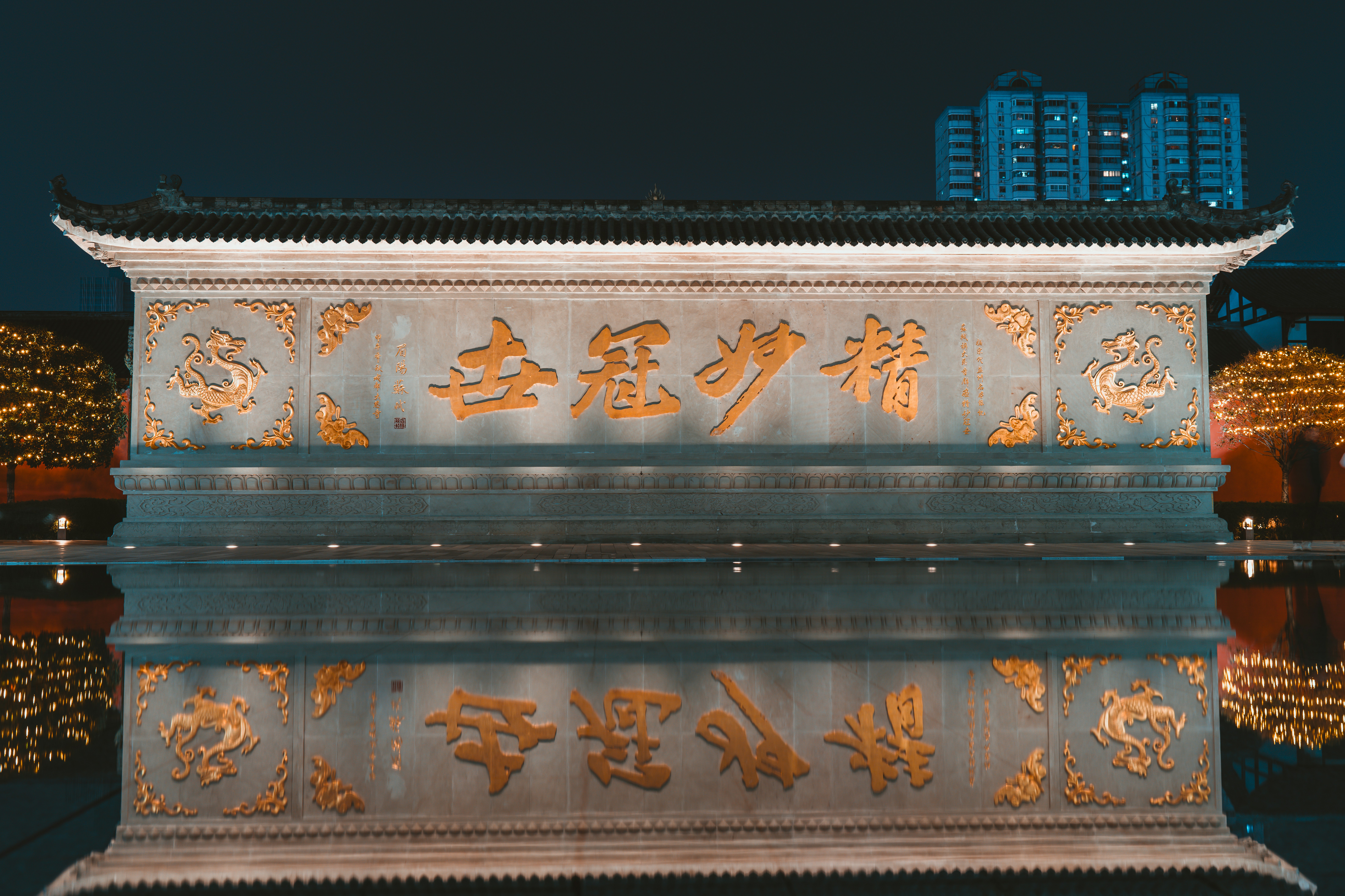 Wall with golden Chinese characters reflected in a calm pool, set against a deep blue night sky.