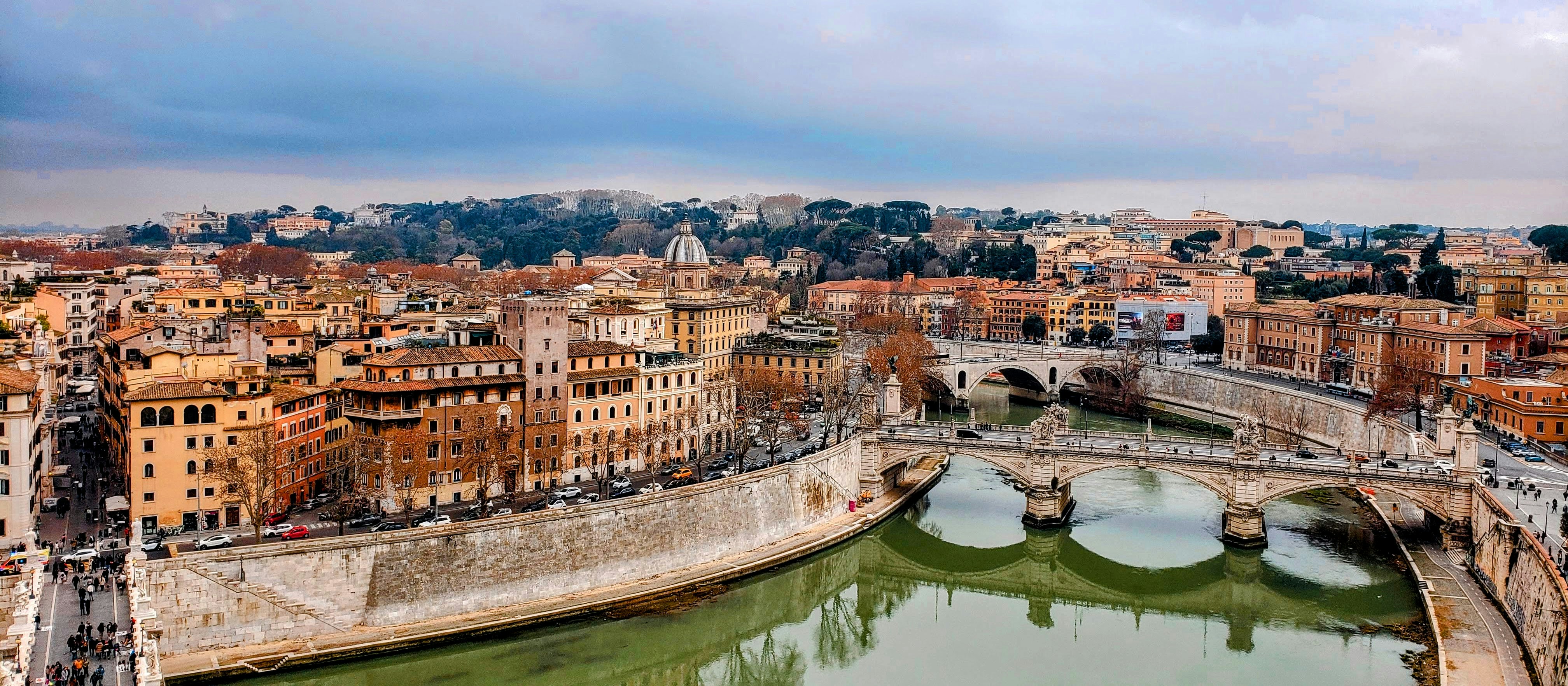 Panoramic view of Rome's historic architecture along the Tiber River under a cloudy sky.