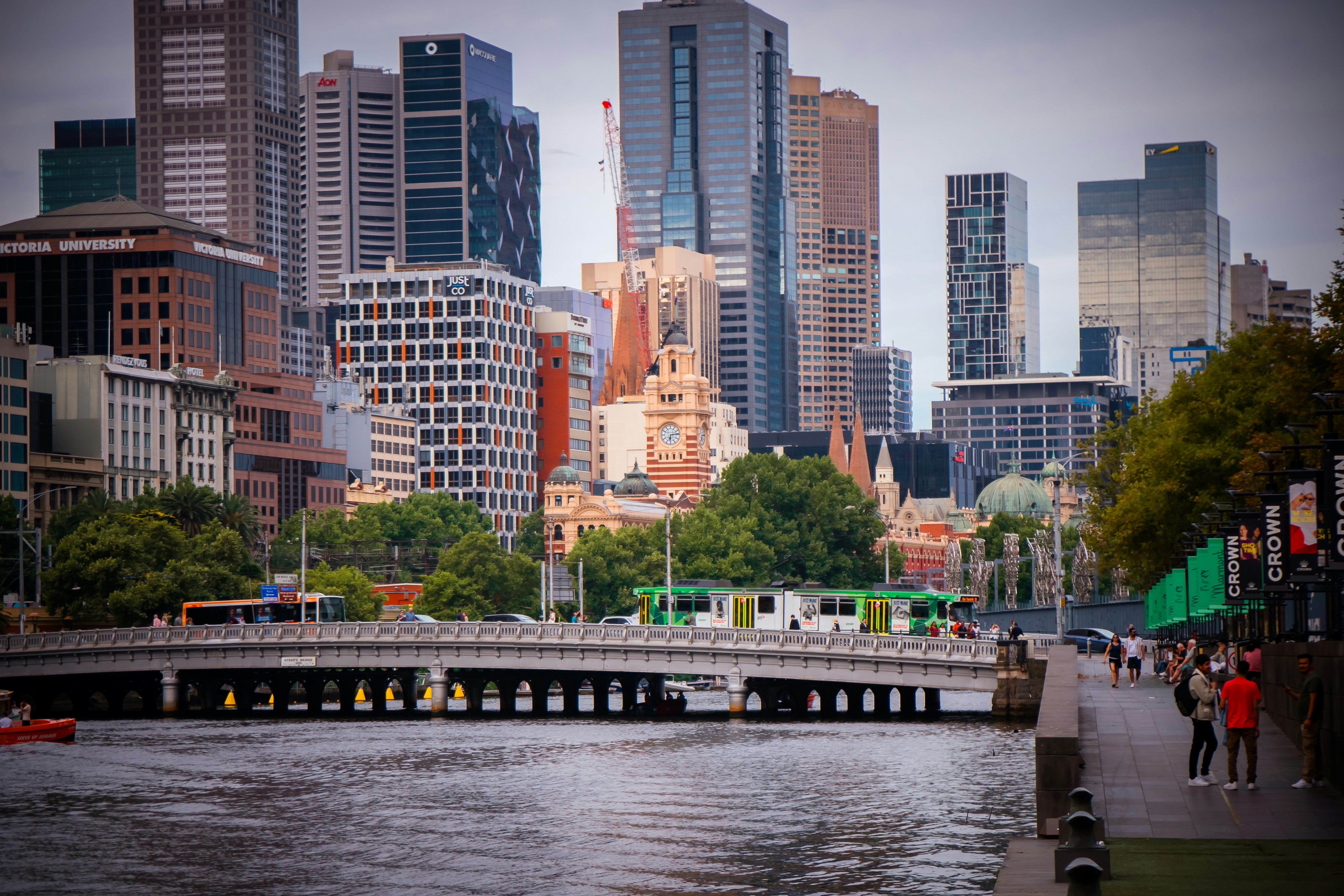 Skyscrapers rise above a tranquil riverbank with walking paths and lush greenery.