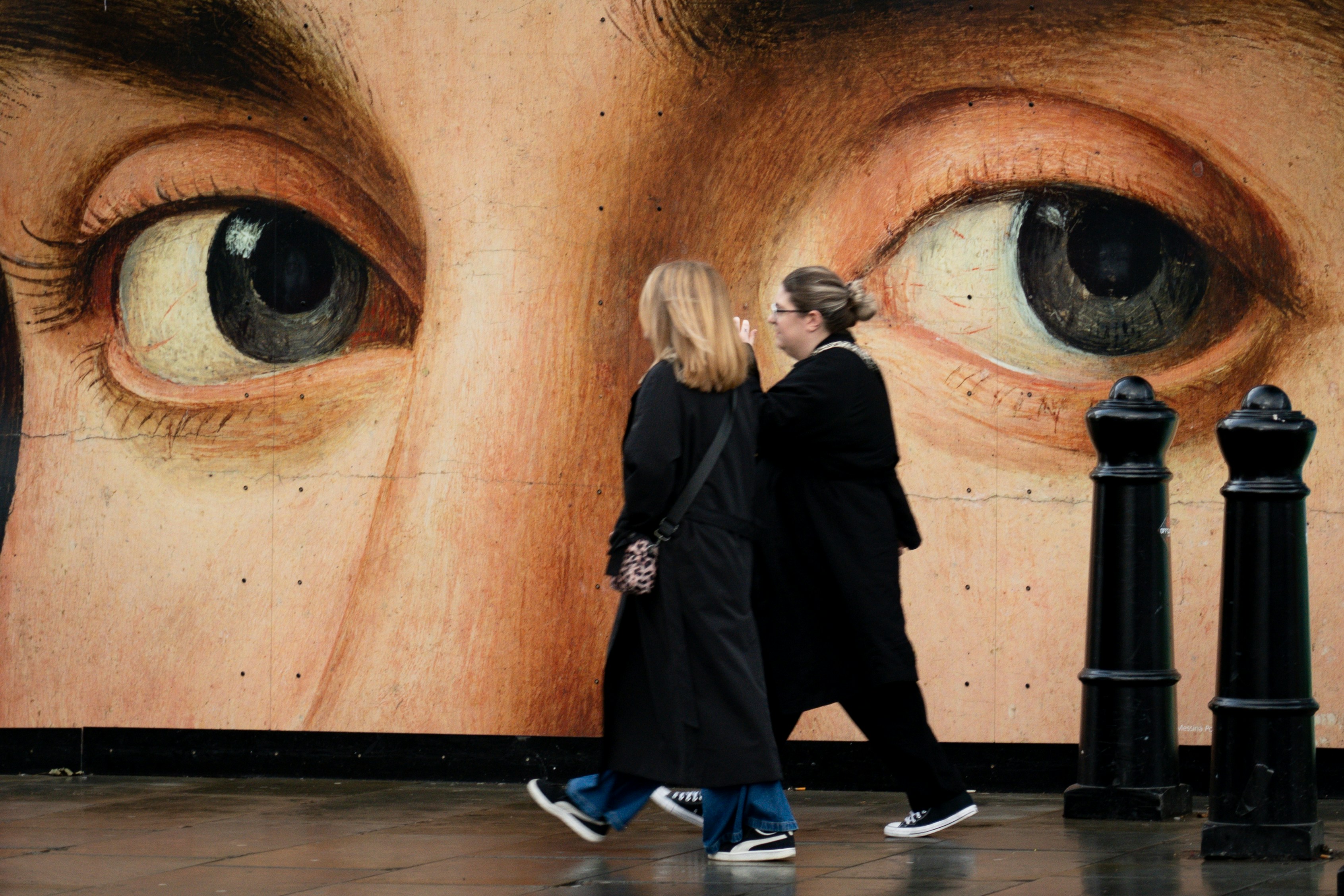 Two individuals walking past a mural of large eyes, with muted clothing contrasting the vibrant artwork.