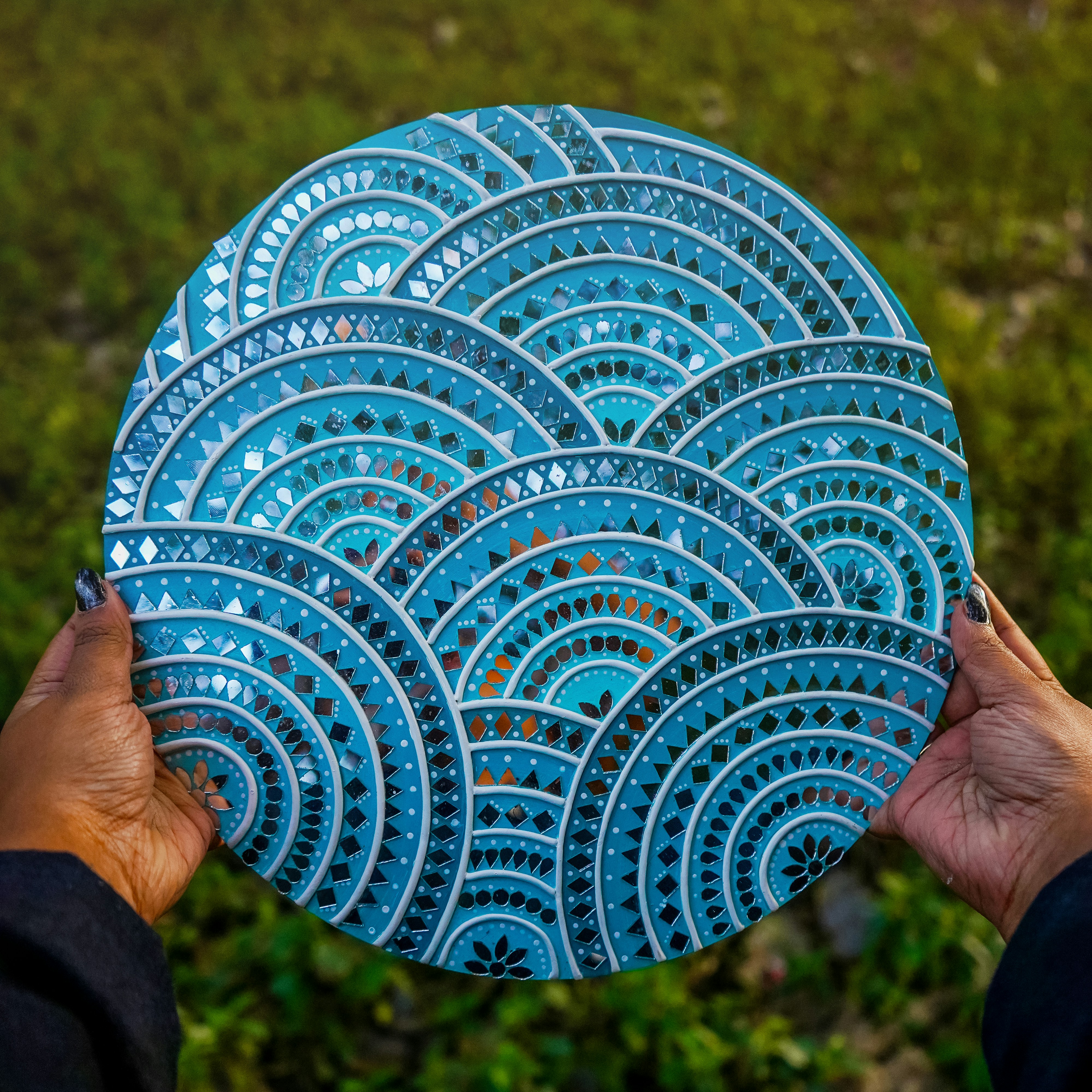 A ceramic plate with an intricate blue mandala pattern is held by two hands, set against a softly blurred grassy background.