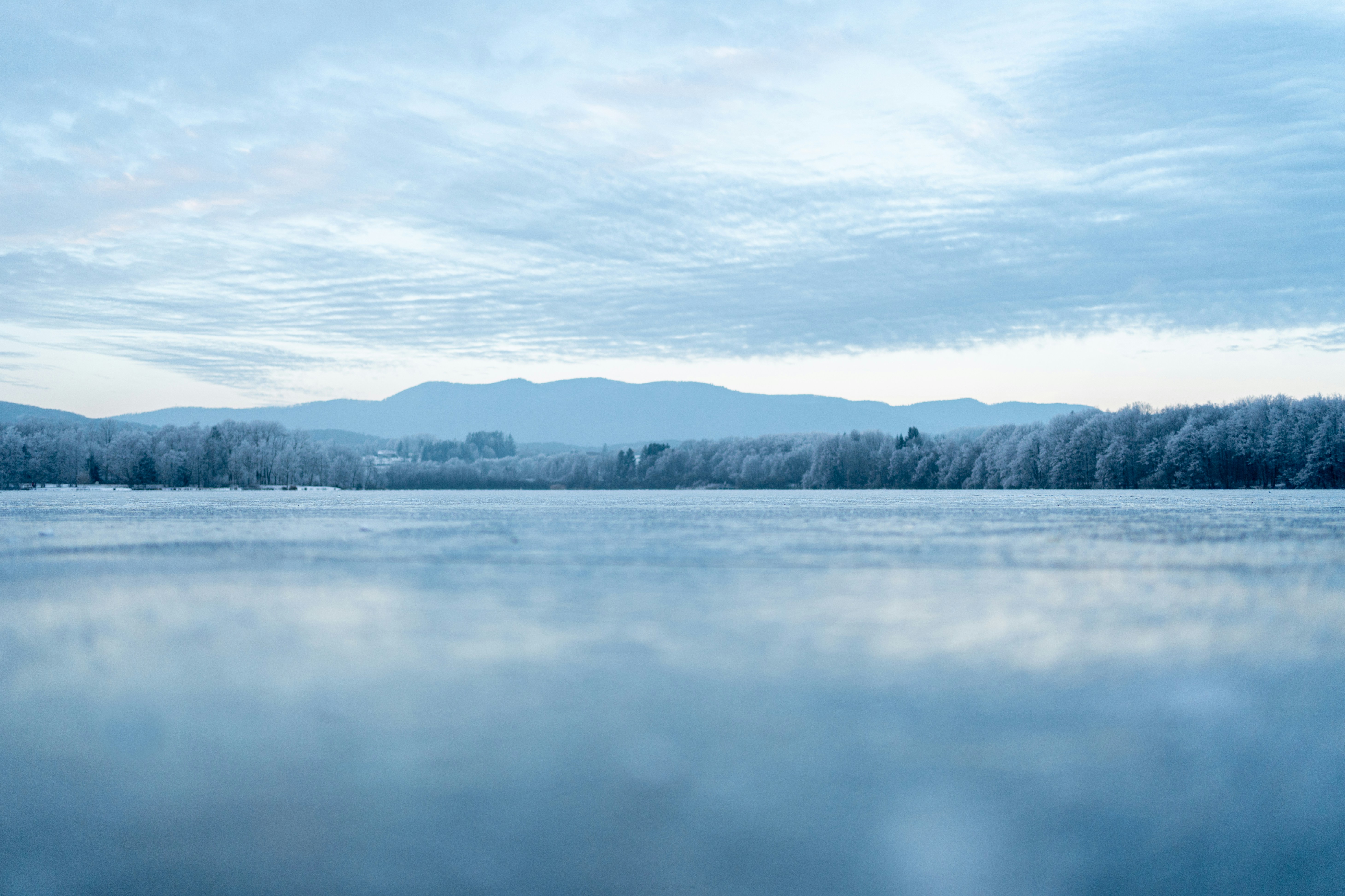 Frozen lake reflecting a serene sky with distant mountains and frosty trees at sunrise.