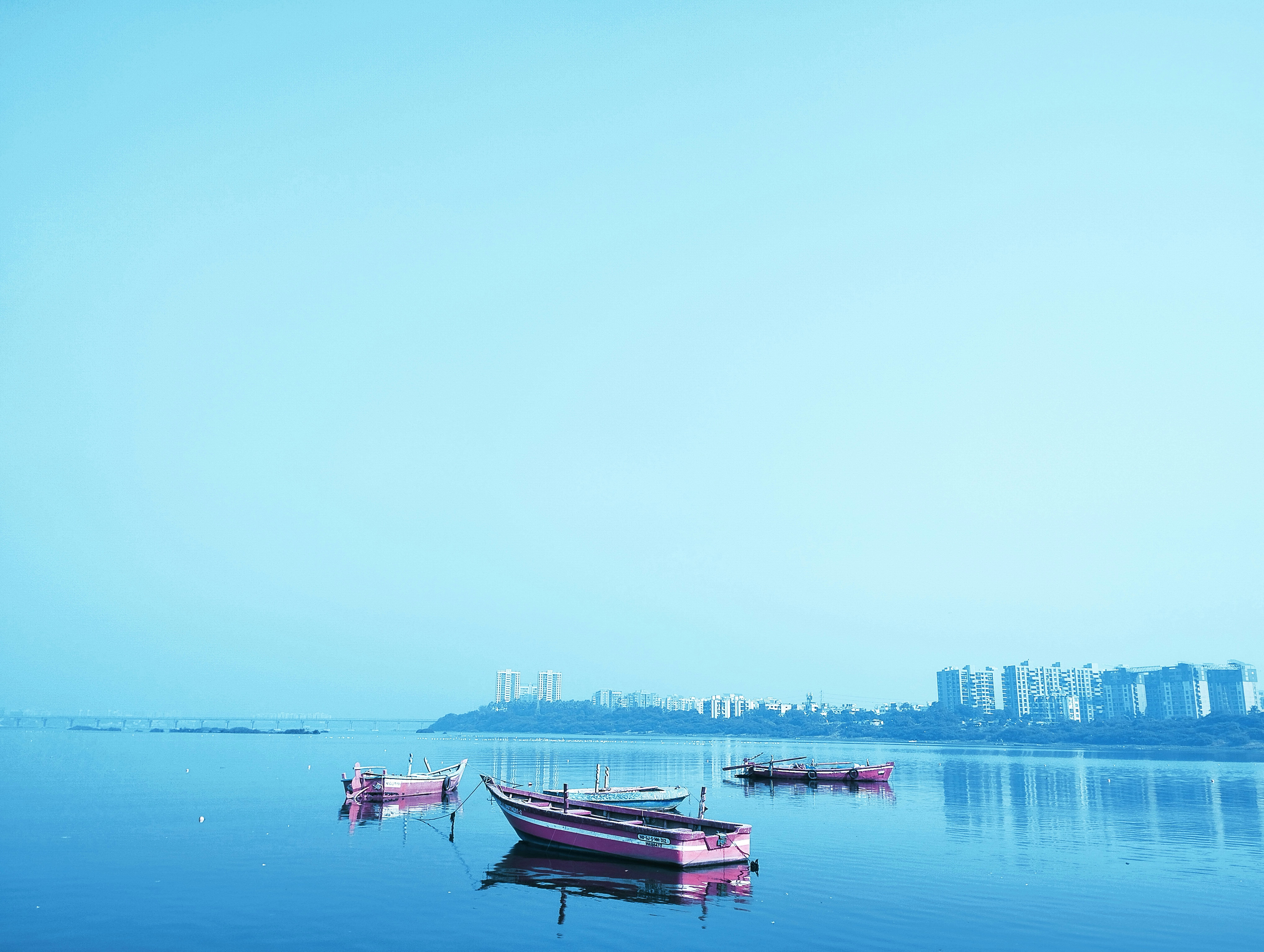Small boats rest on a calm river against a distant city skyline under a clear blue sky.