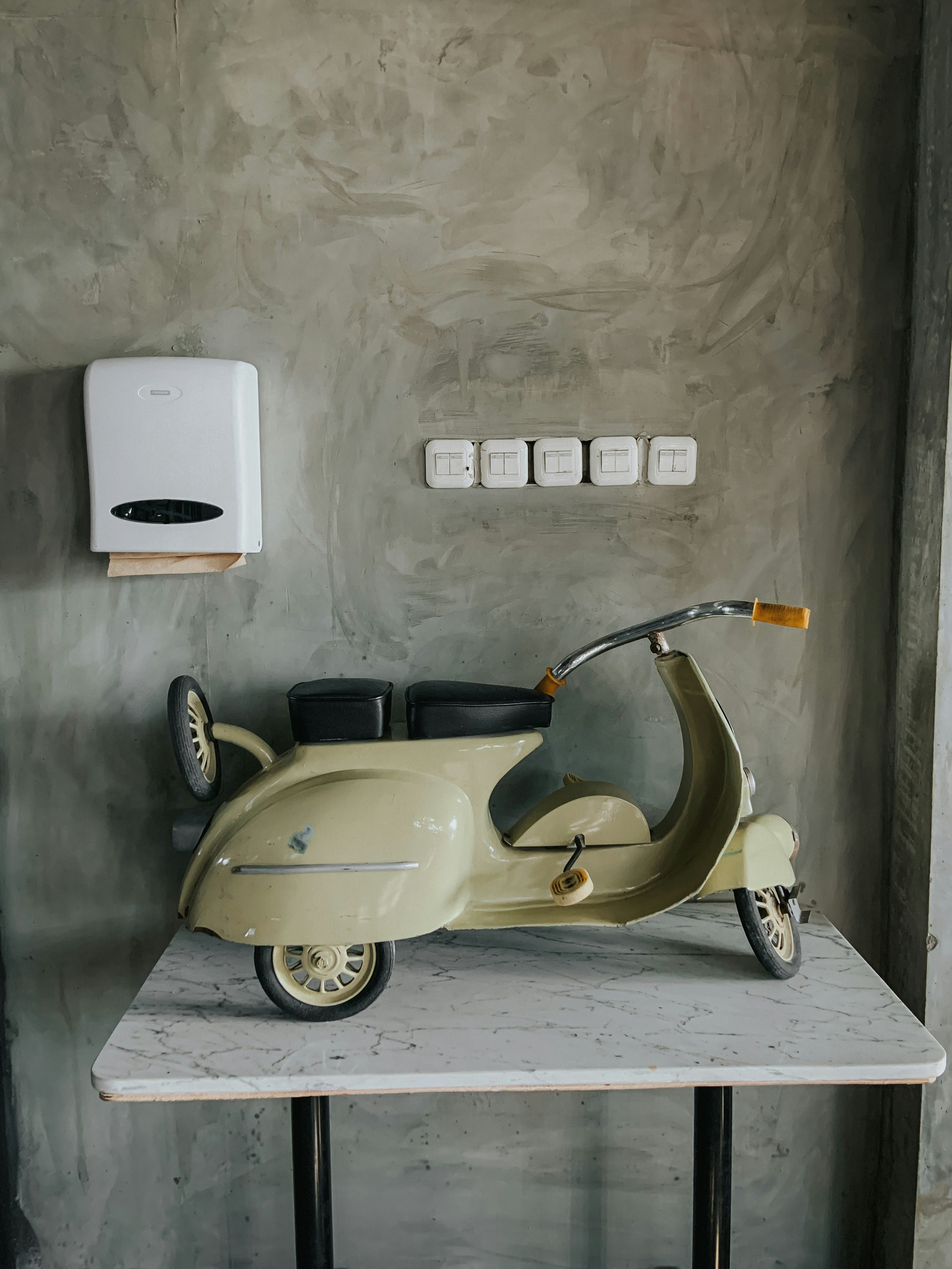 Beige Vespa-Style Toy Scooter on Marble Table Against a Textured Concrete Wall