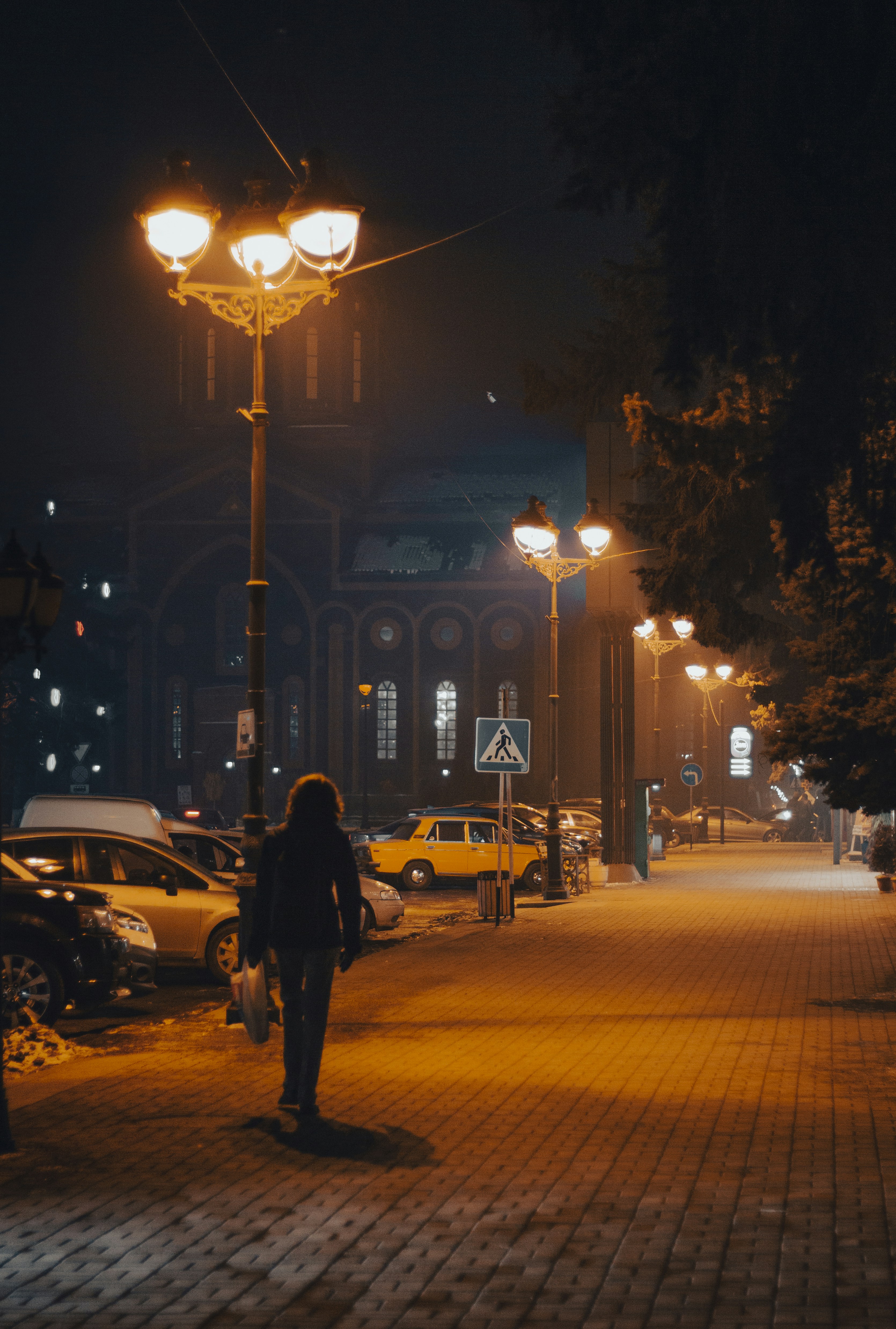 Person walking along a dimly lit street lined with parked cars and glowing streetlamps at night.