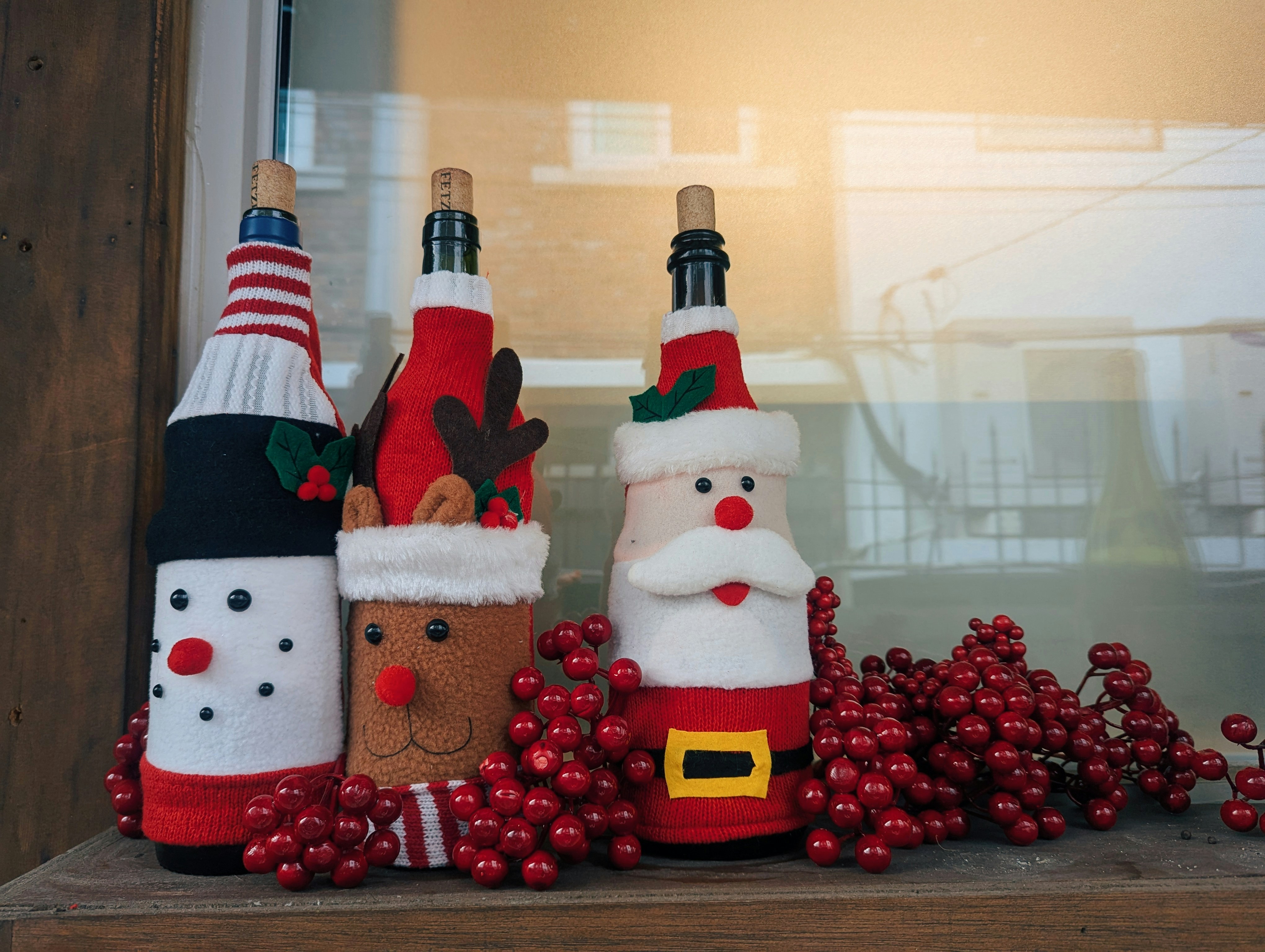 Three wine bottles adorned with holiday-themed covers, surrounded by clusters of red berries.