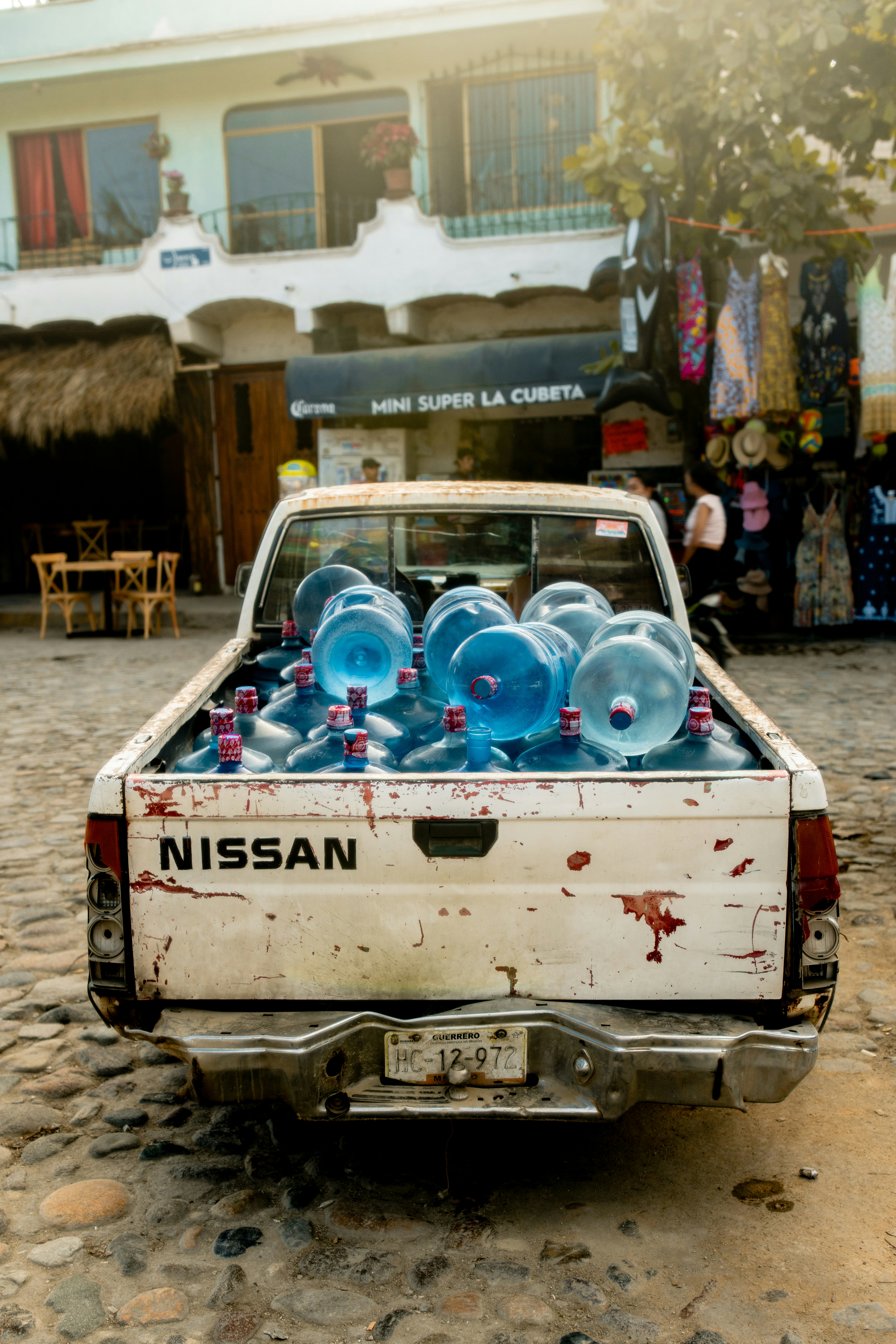 Blue Jug Load in a Weathered Nissan: Sunlit Cobblestones and Market Backdrop