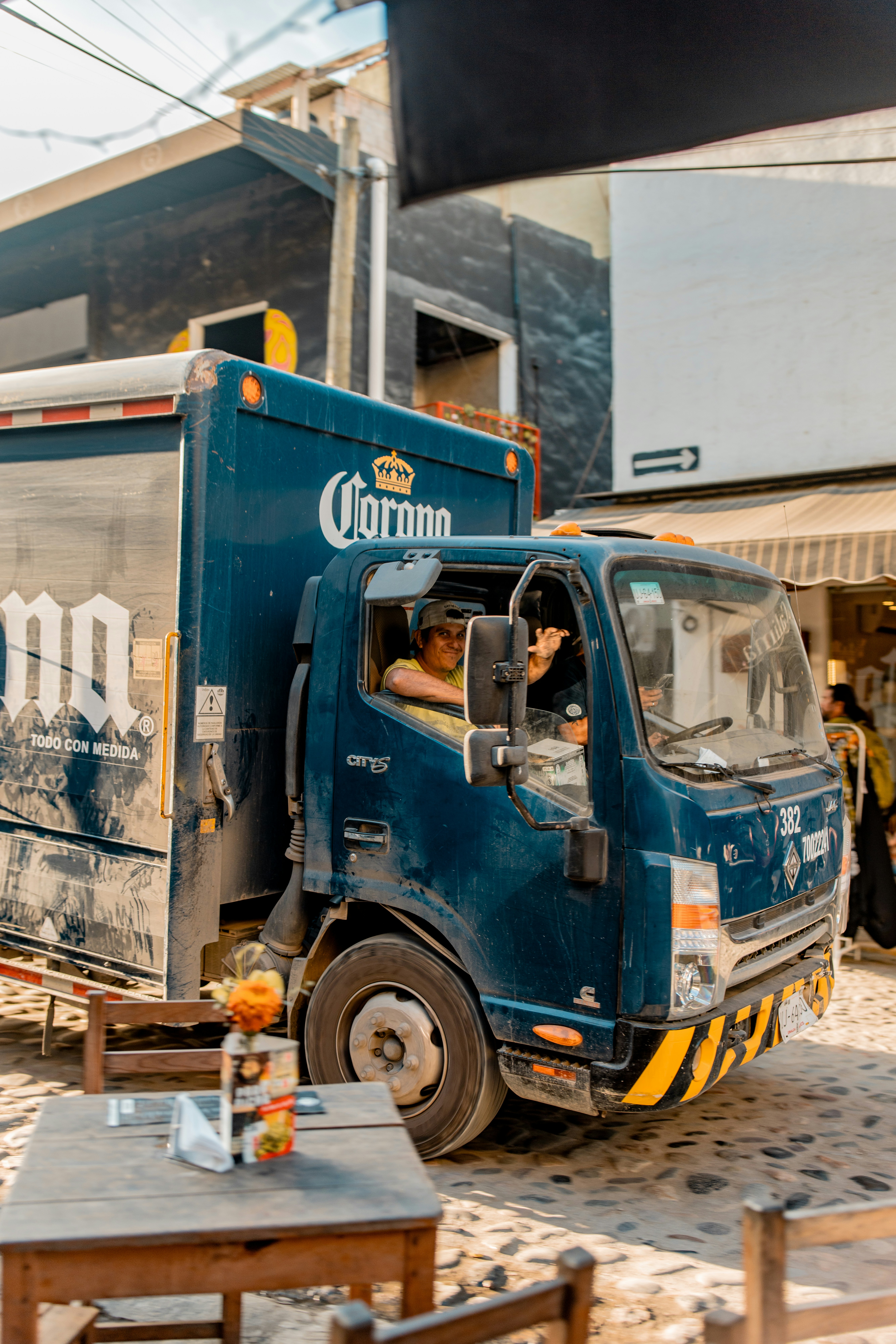 Blue Corona Truck Pauses at a Sunlit Cobblestone Street Market