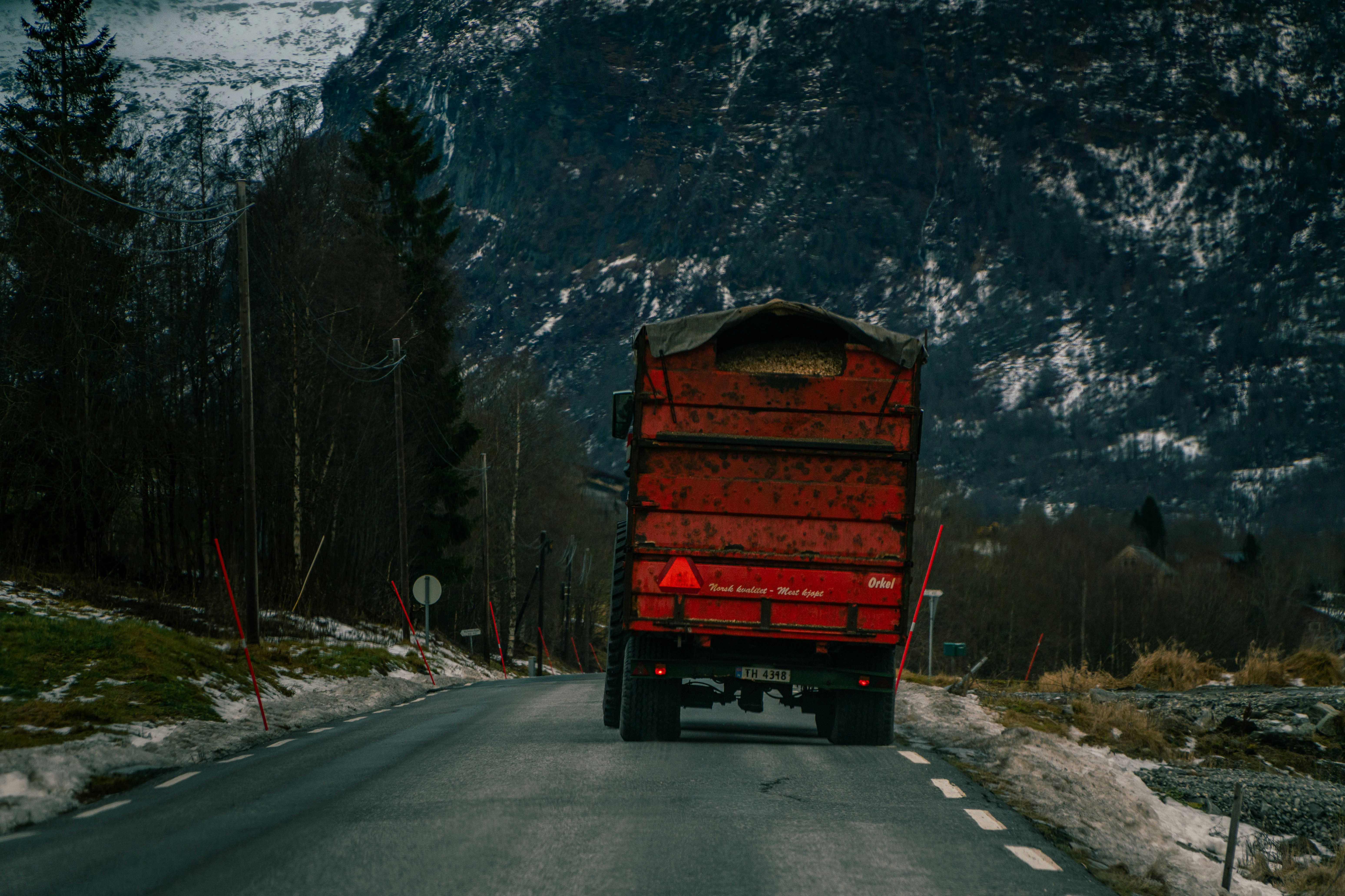 A red farm truck full of grain is pulled by a tractor along a mountain pass.