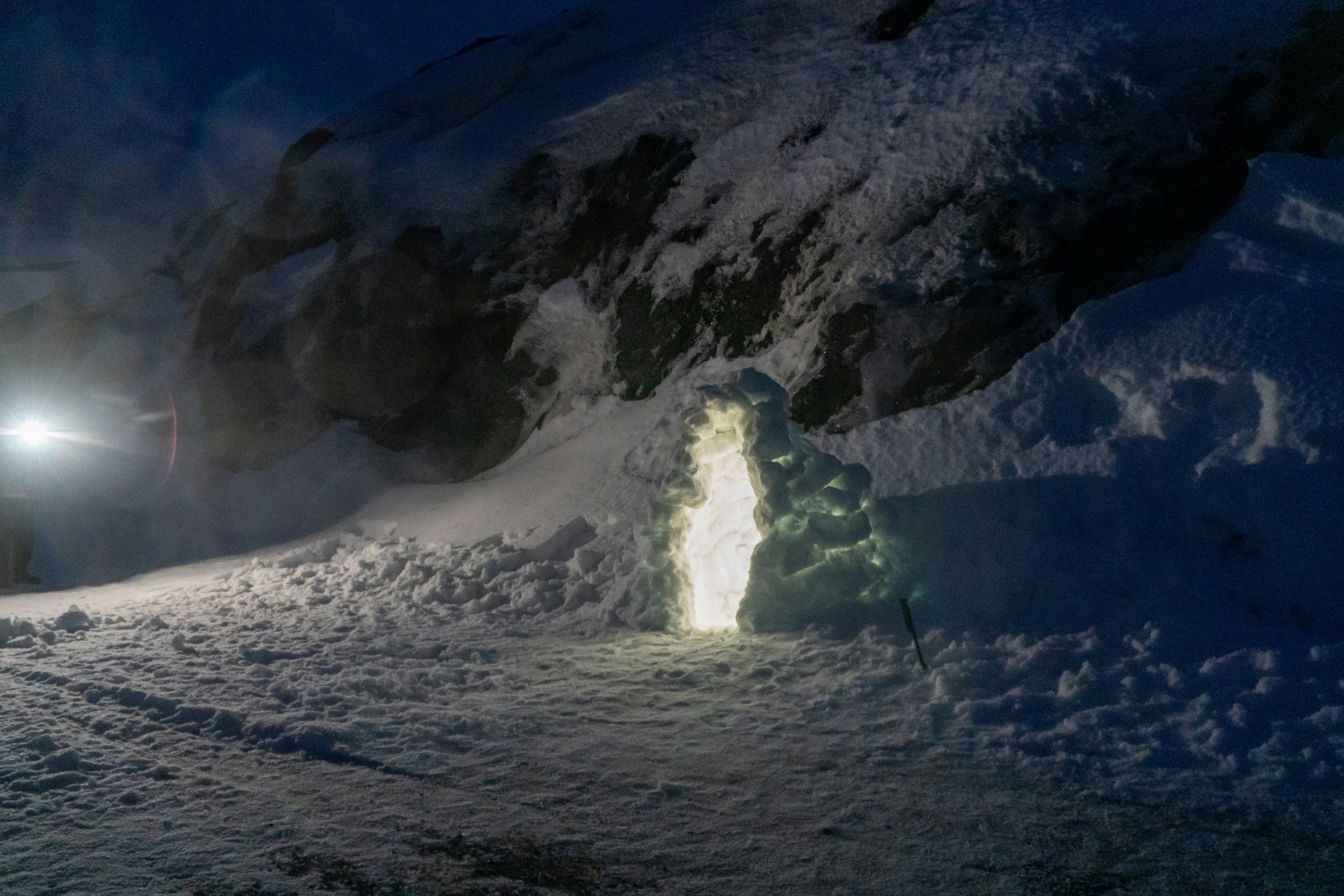 Snow cave with a warm light glowing inside, contrasting against the dark, snowy landscape at dusk.