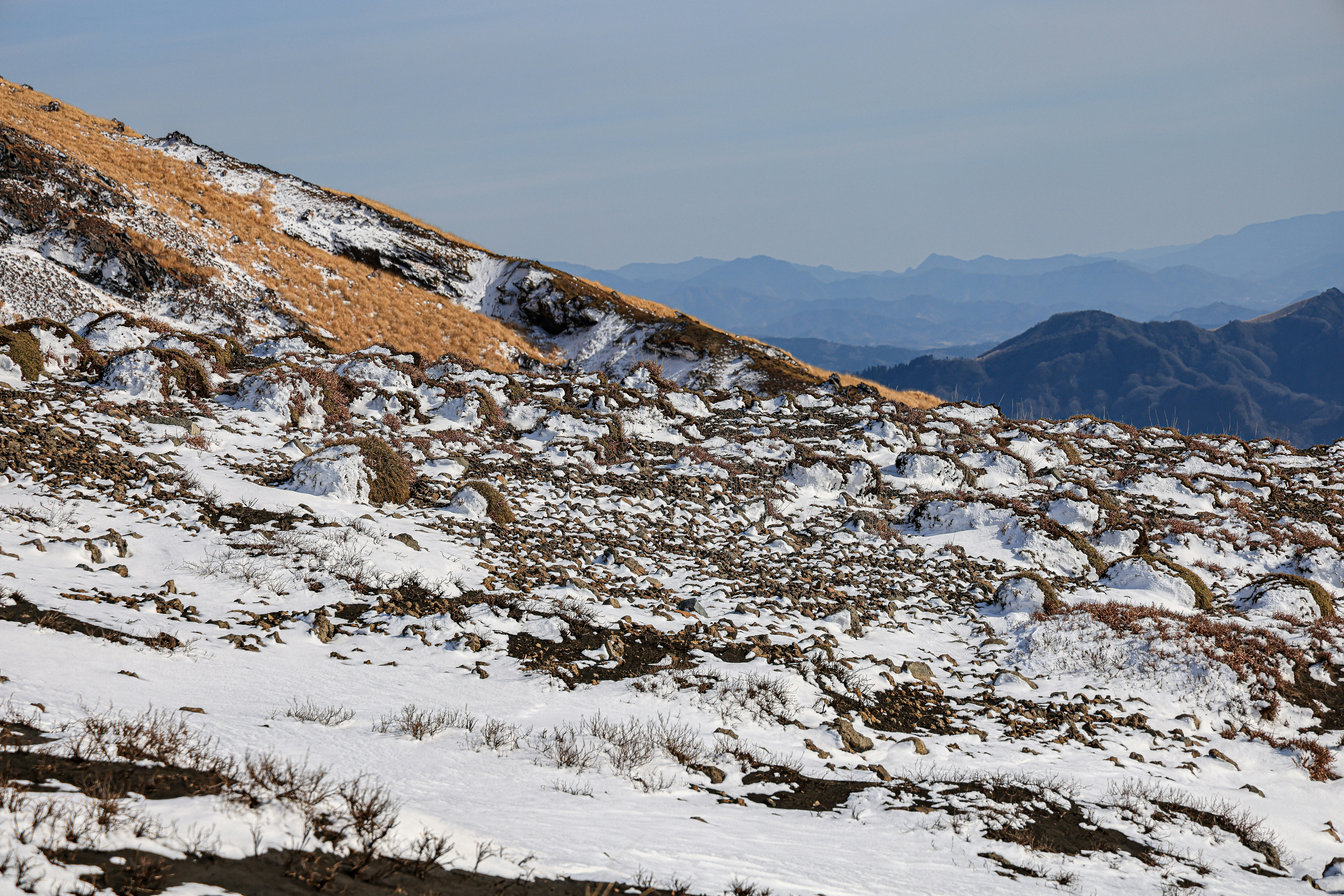 Snow-dusted rocky hillside with golden grass and distant mountain range under a clear sky.