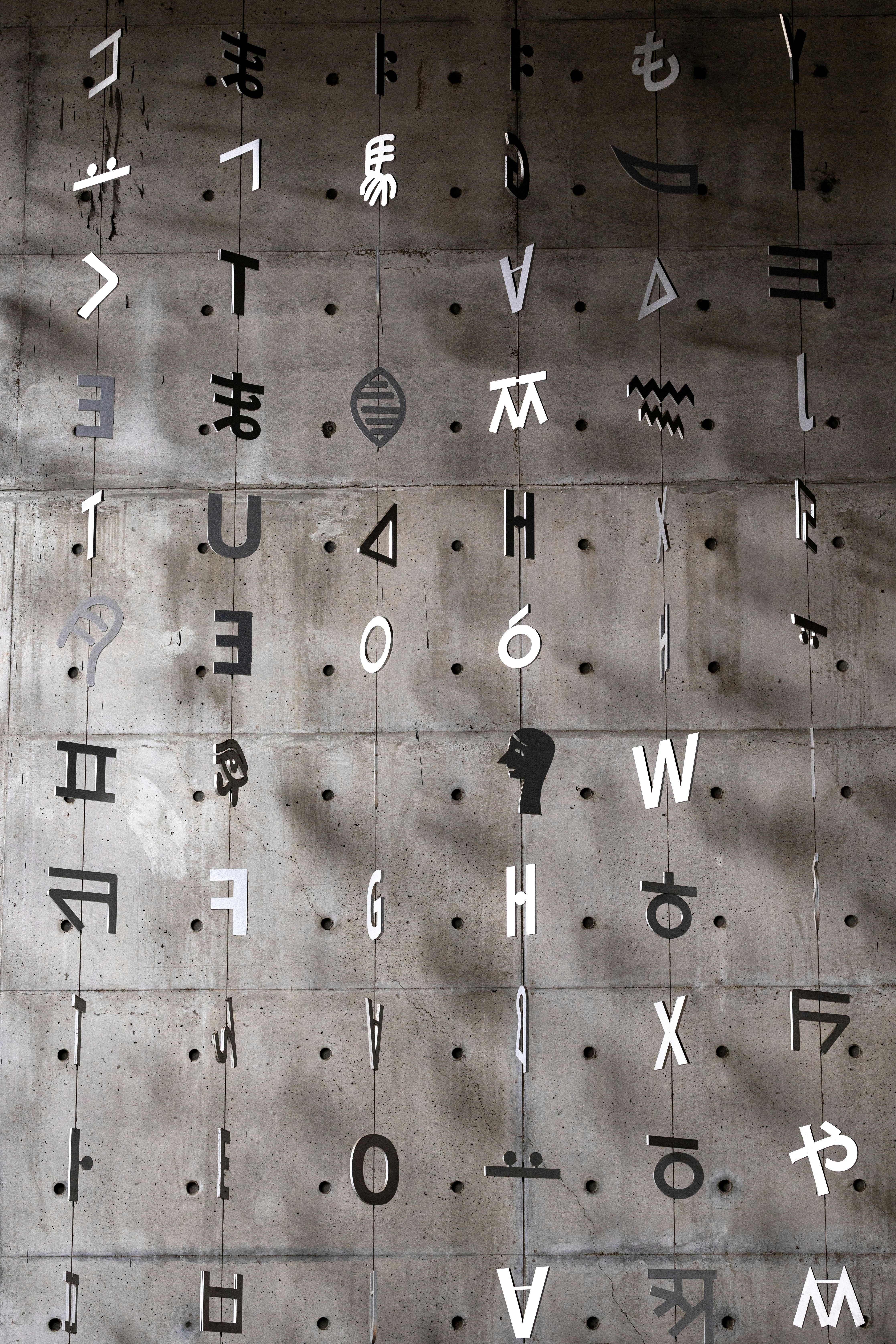 Photograph of a tiled concrete wall segmented into panels with white symbols and letters. A dark silhouette resembling a head sits along a vertical line near the center.