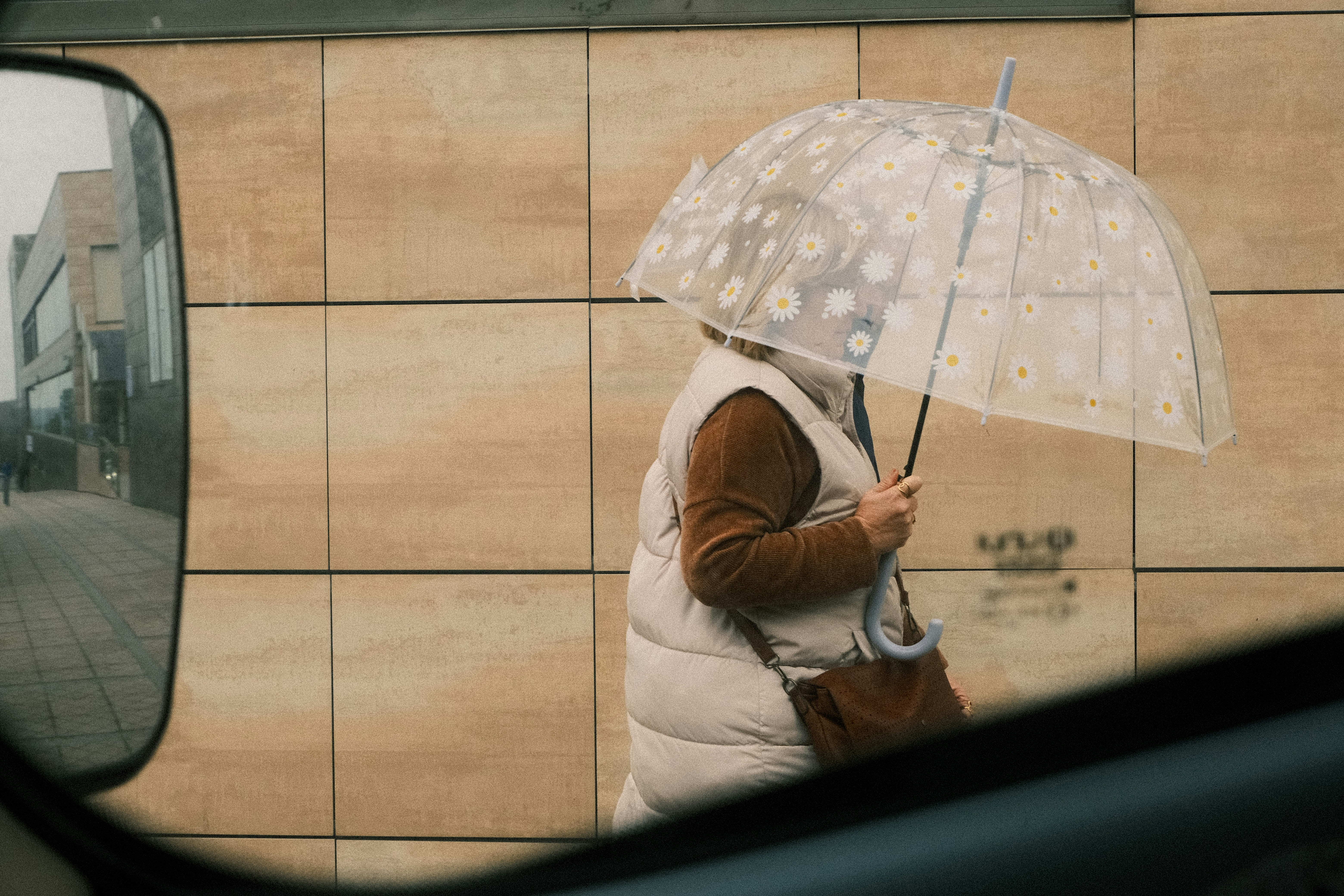 Person with a polka-dotted umbrella strolling past a tiled wall on a quiet street.
