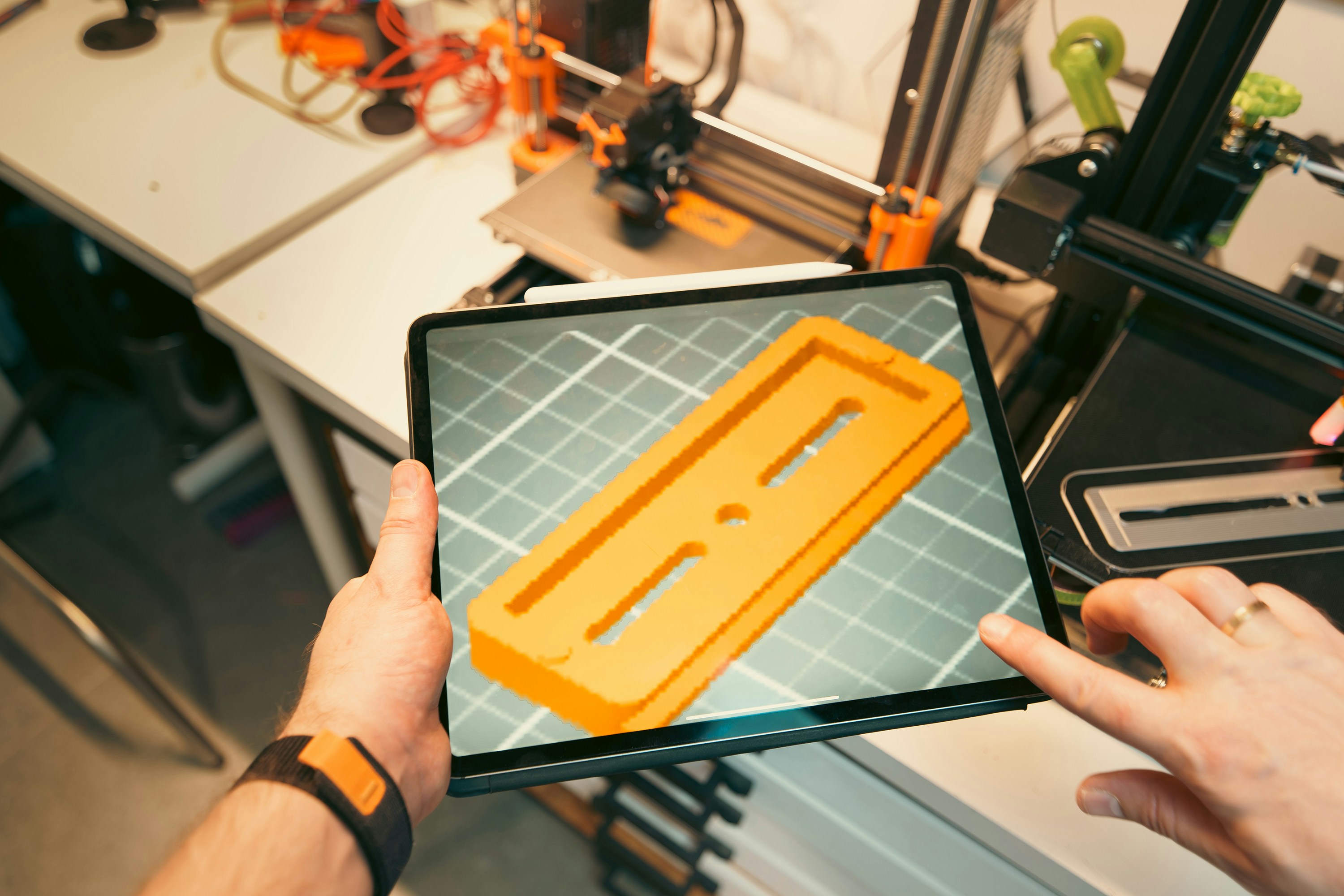Hands holding a tablet displaying an orange 3D model against a grid, with a 3D printer in the background.