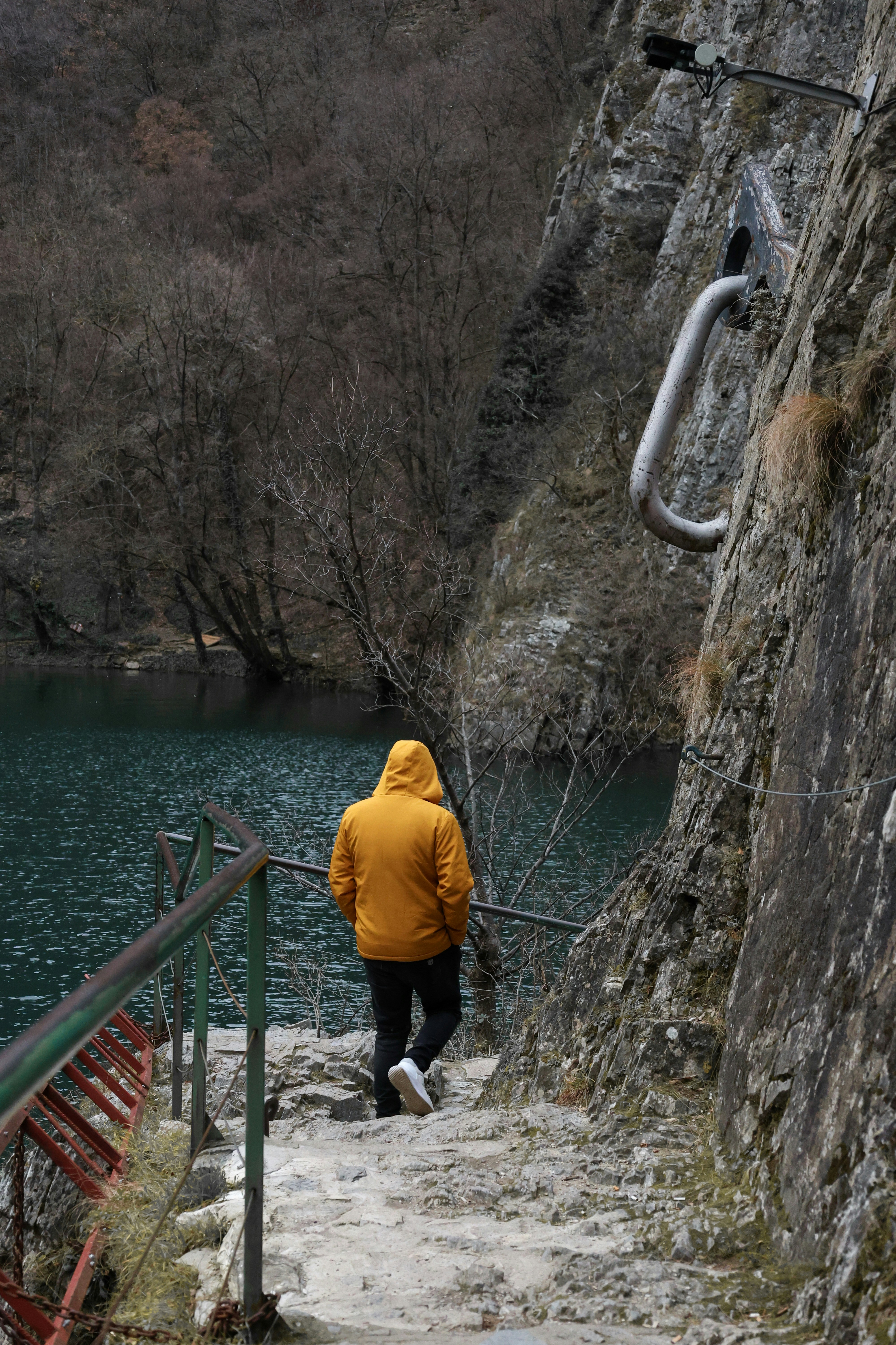 Amber Hooded Traveler Navigates a Narrow Cliffside Staircase Beside Teal Lake