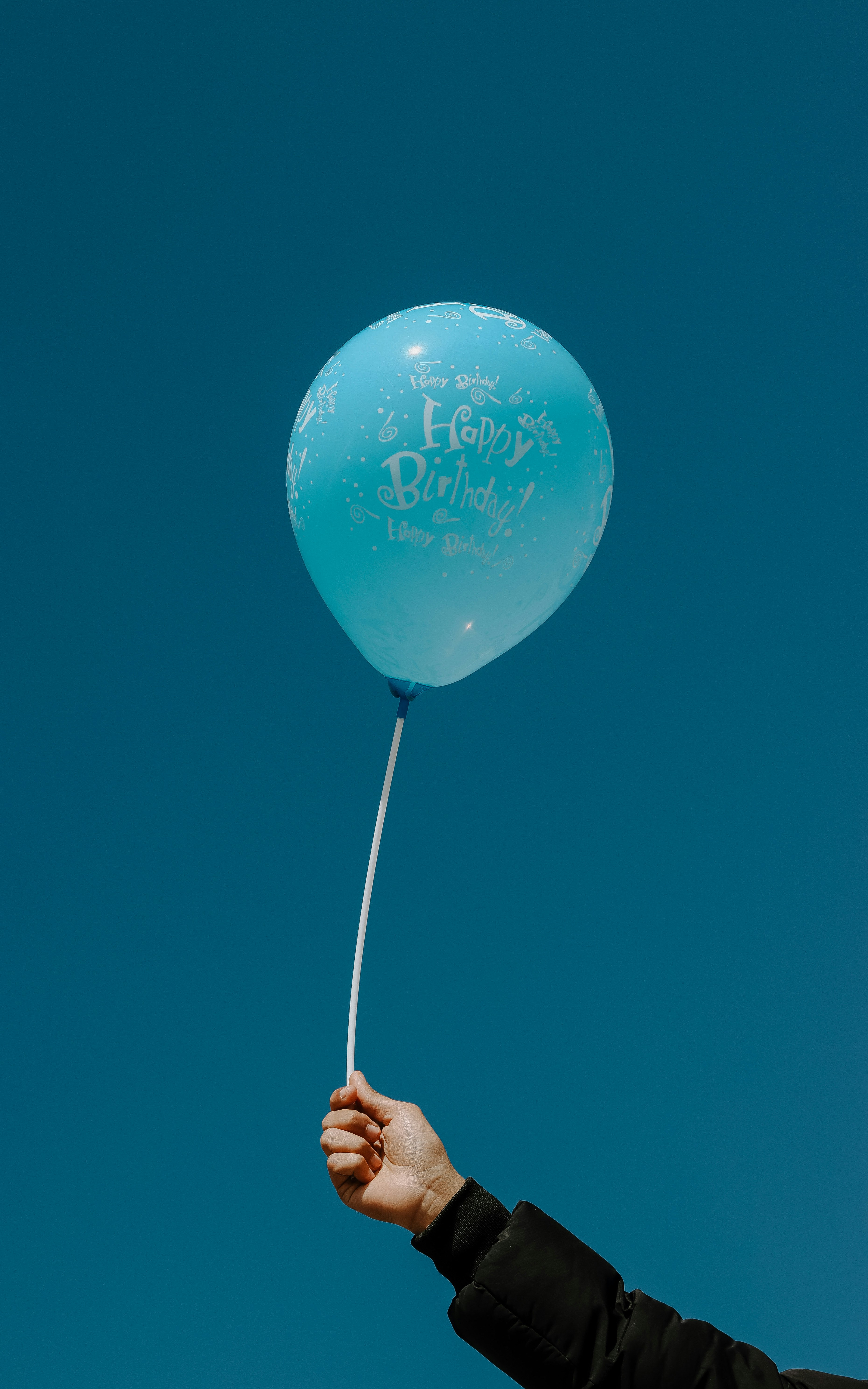 Blue birthday balloon tethered by a hand rises against a deep blue sky.
