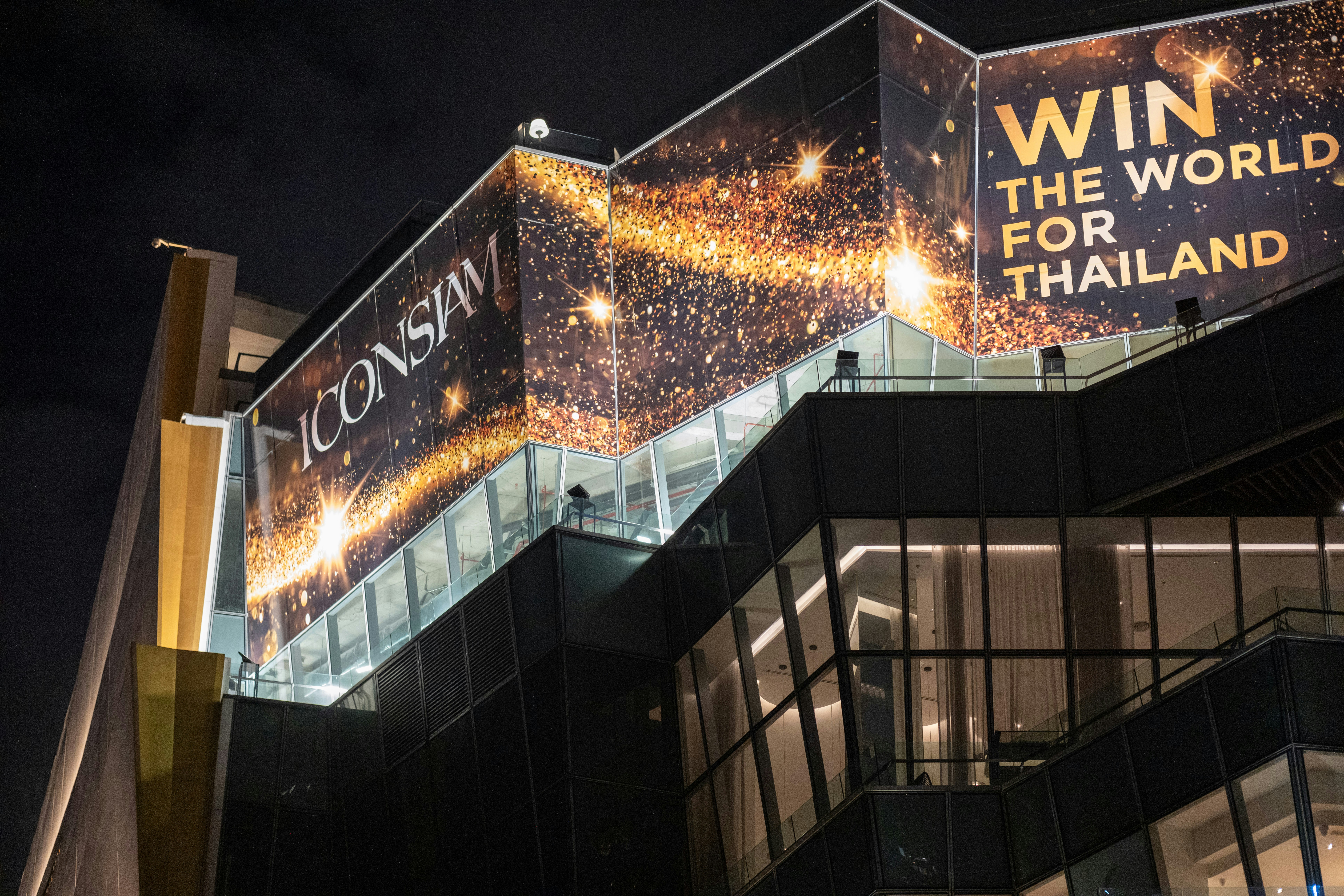 ICONSIAM's facade glows with golden lights against a dark sky, showcasing modern glass and steel architecture.