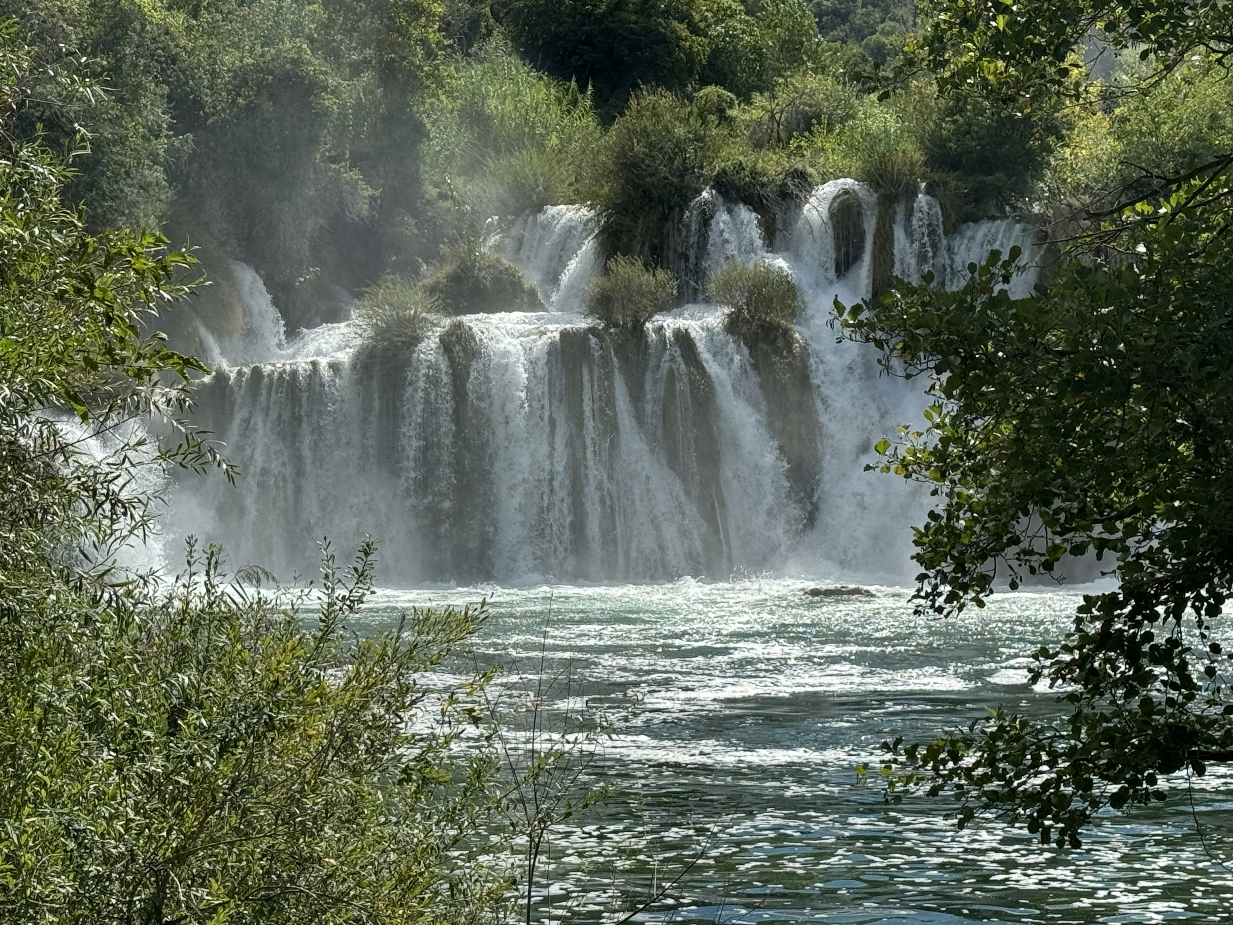 Waterfall cascading over rocky ledges surrounded by dense green foliage, shimmering in dappled sunlight.