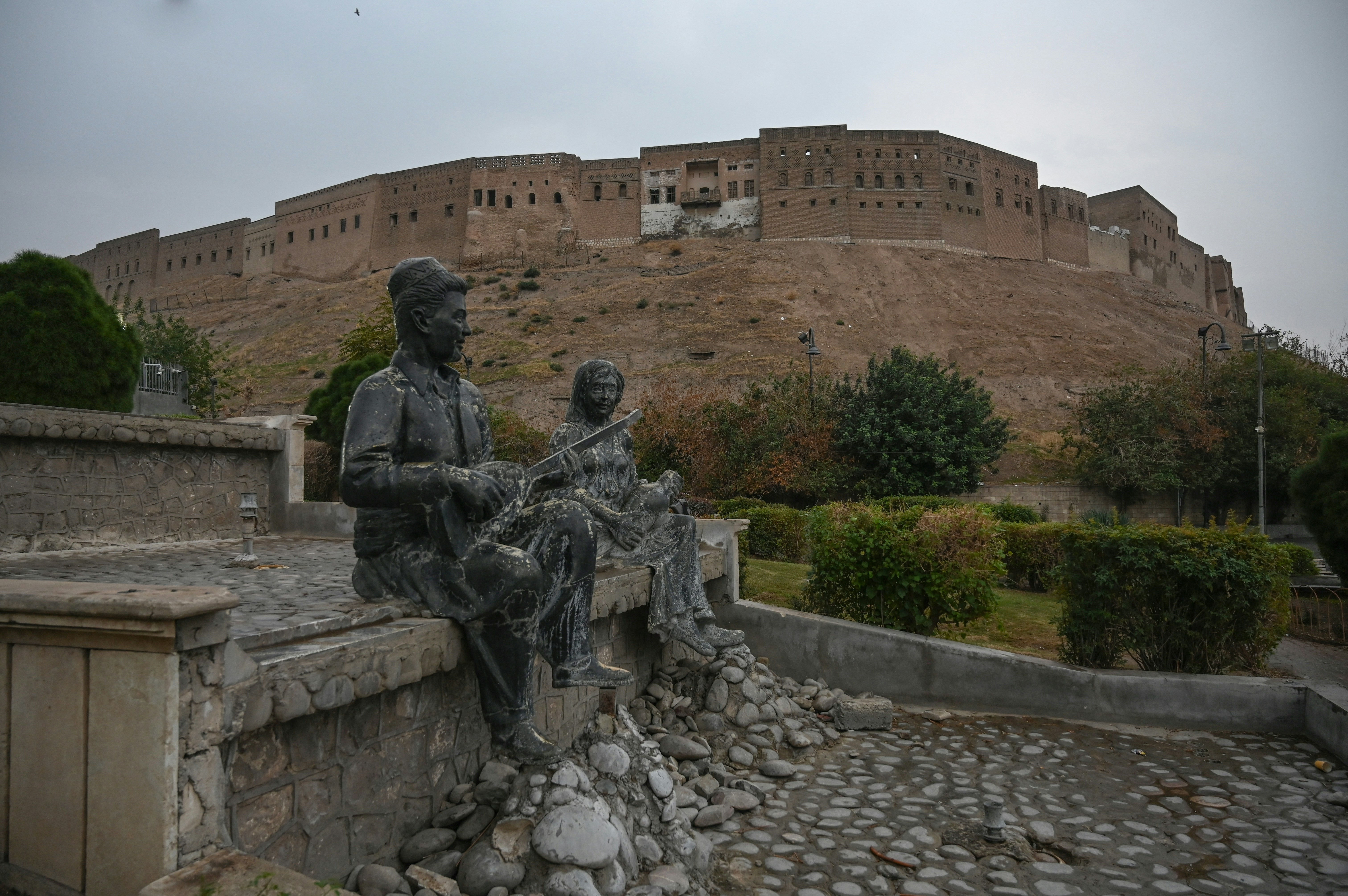 Two statues playing instruments with a historic fort towering in the background under an overcast sky.