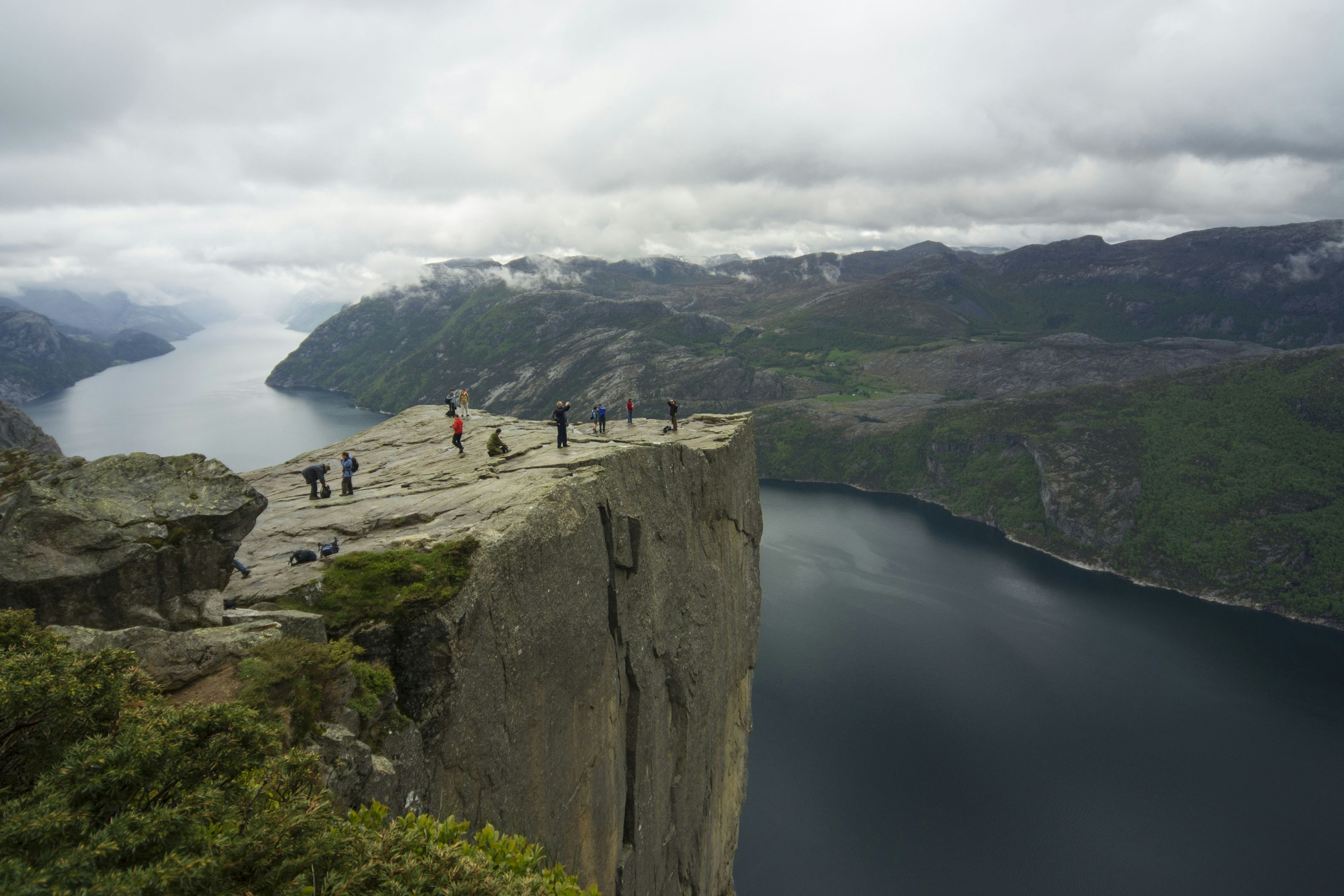 Hikers standing atop a sheer cliff overlooking a serene fjord under a cloudy sky.