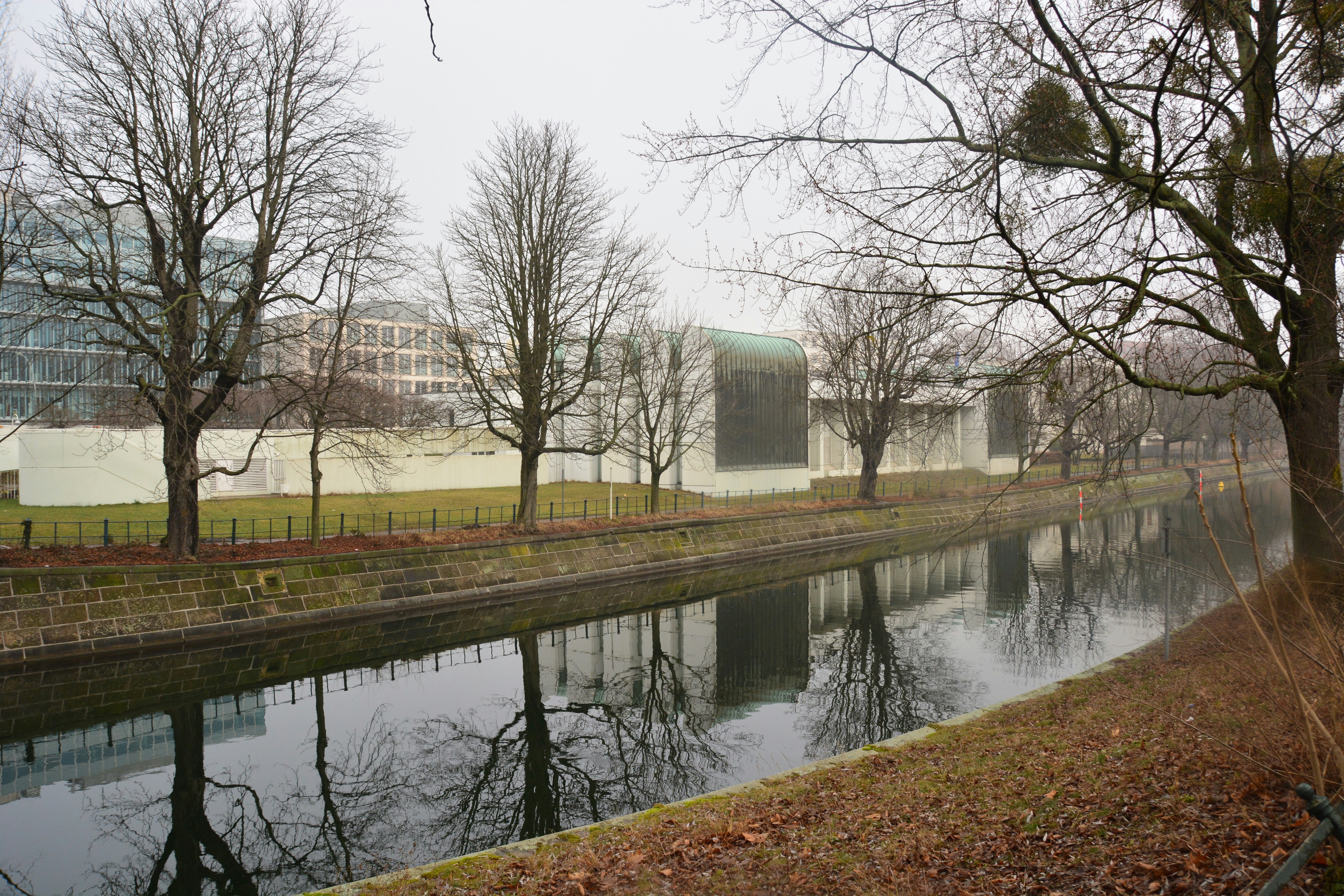 Tranquil canal reflecting bare trees and modern architecture under an overcast sky.
