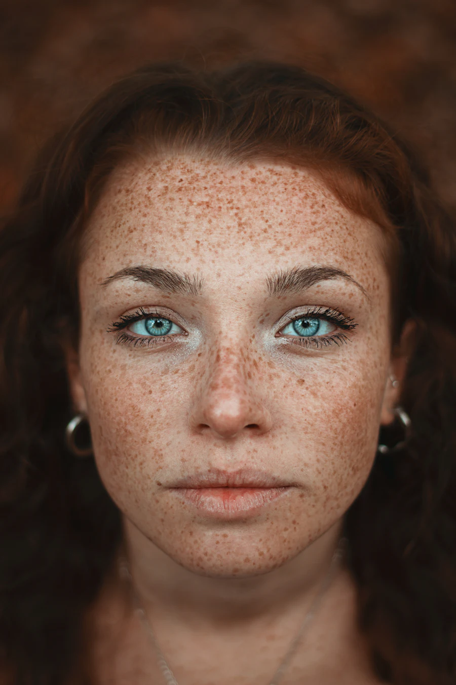 Close-up portrait of a freckled woman with bright blue eyes and dark hair against a softly blurred brown background. Her face is centered and lit evenly, with hoop earrings and a calm expression.