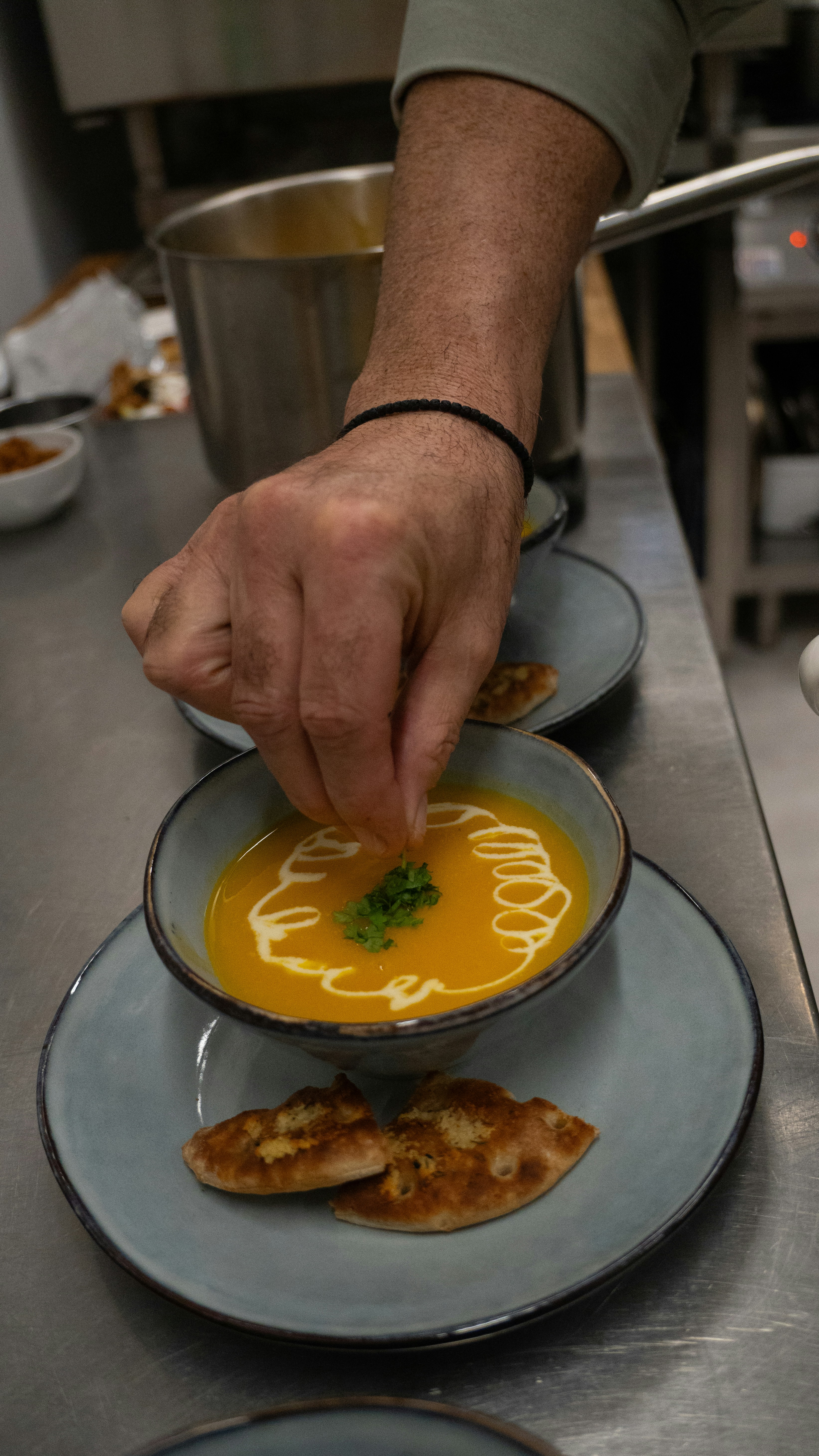 Chef garnishing a vibrant bowl of soup with fresh herbs, accompanied by toasted flatbreads on a sleek countertop.