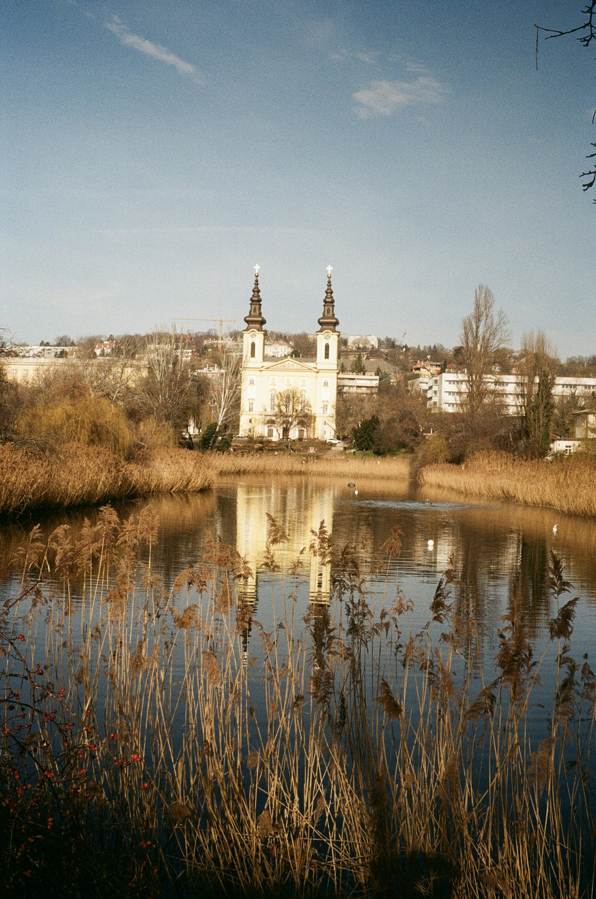 Twin-spired church rises over a calm river, its reflection mirrored in the water. Reeds frame the foreground.