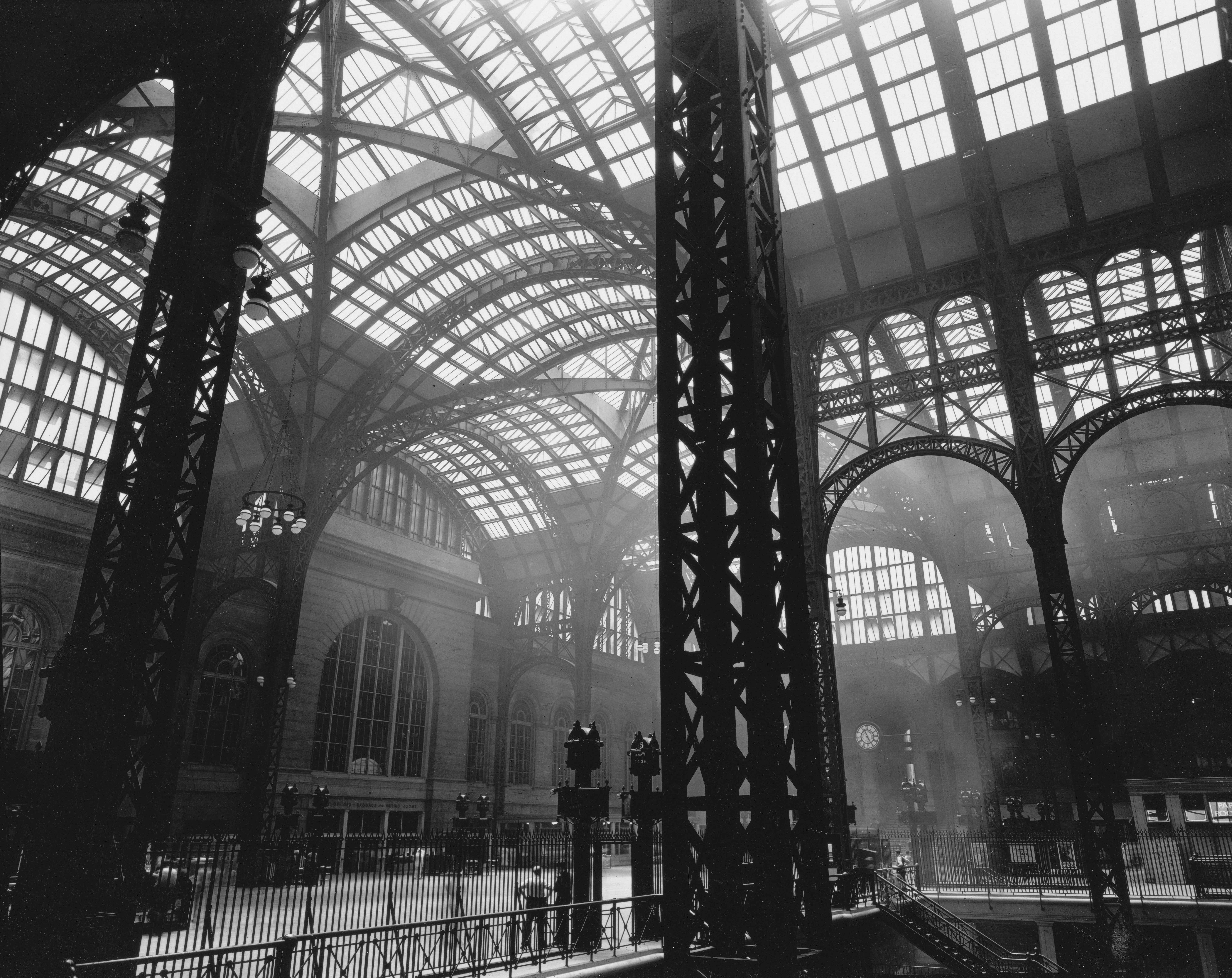 Sunlight streams through the iron arches and glass ceiling of Penn Station's vast interior, highlighting intricate details of early 20th-century architecture.