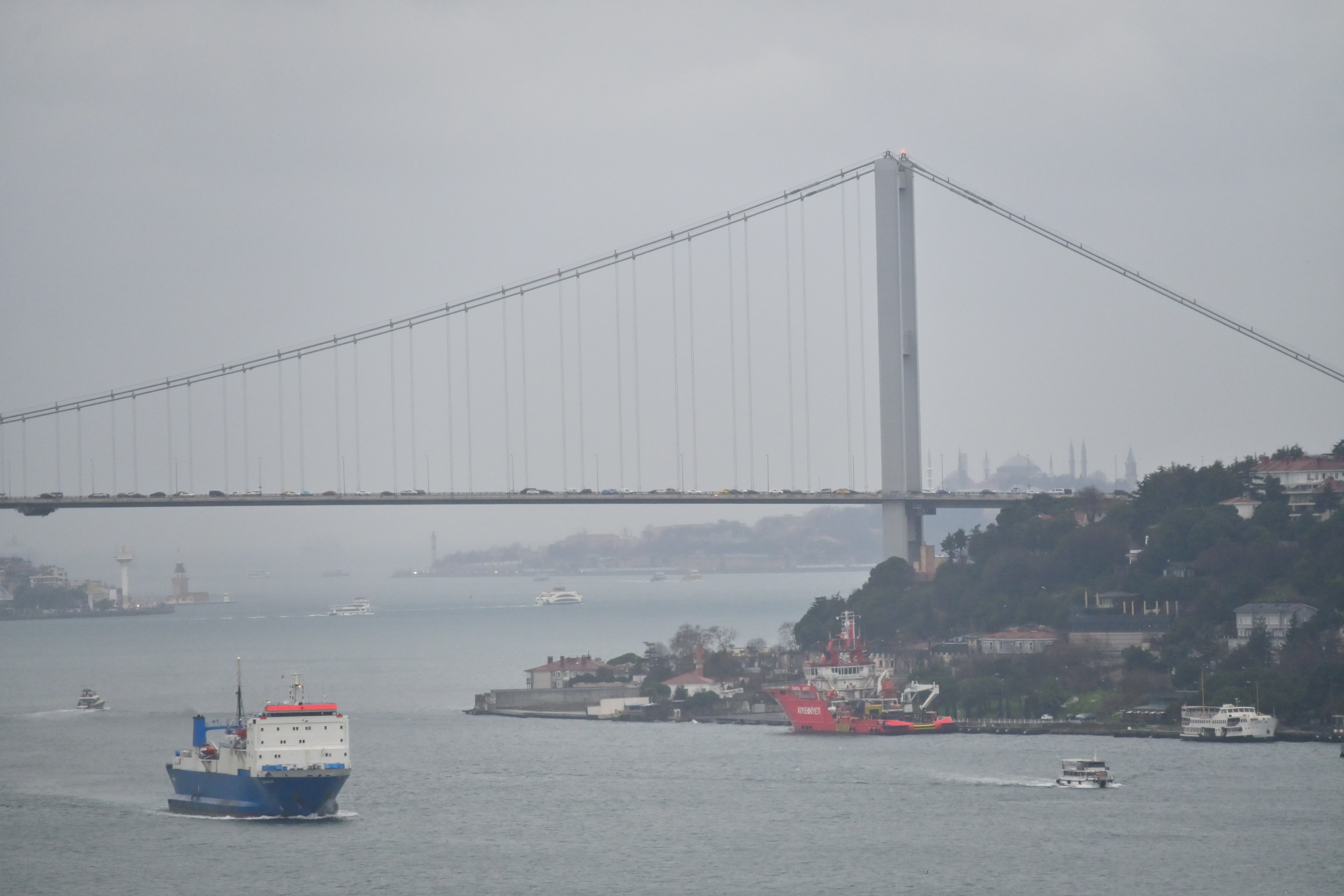 Cargo ship navigating the Bosphorus Strait beneath a towering suspension bridge on a foggy day.