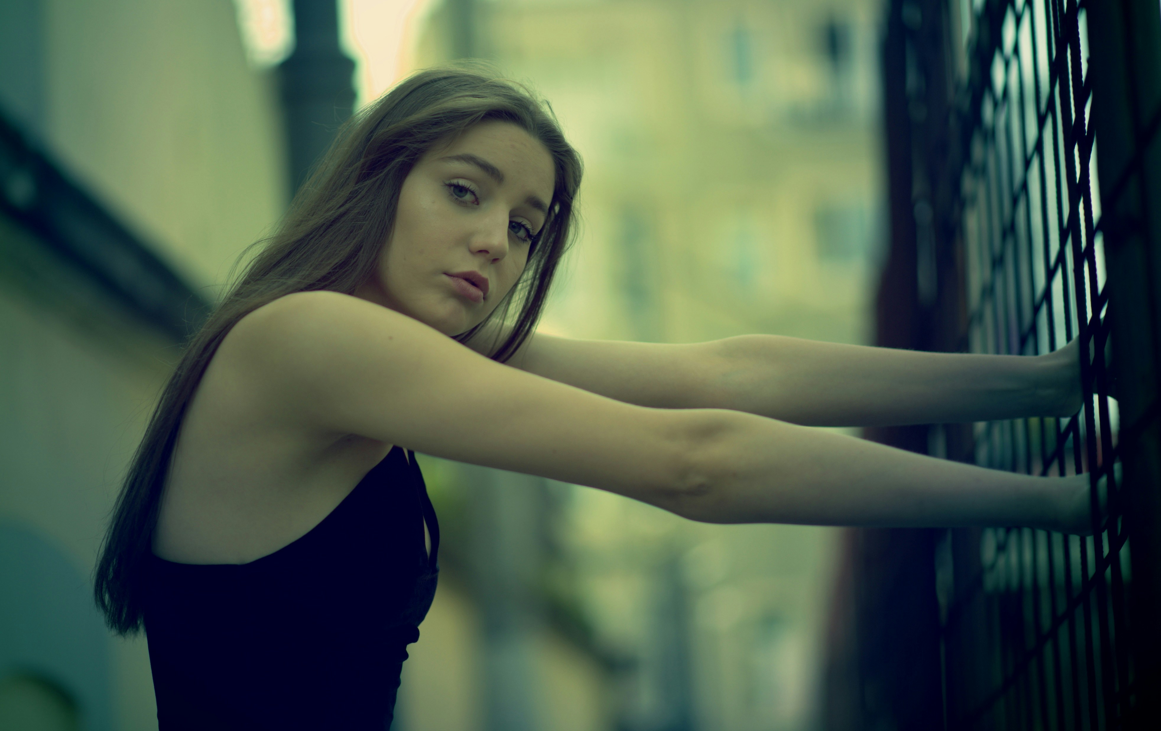 Young woman leans against a fence, lost in thought, with a softly blurred urban background.