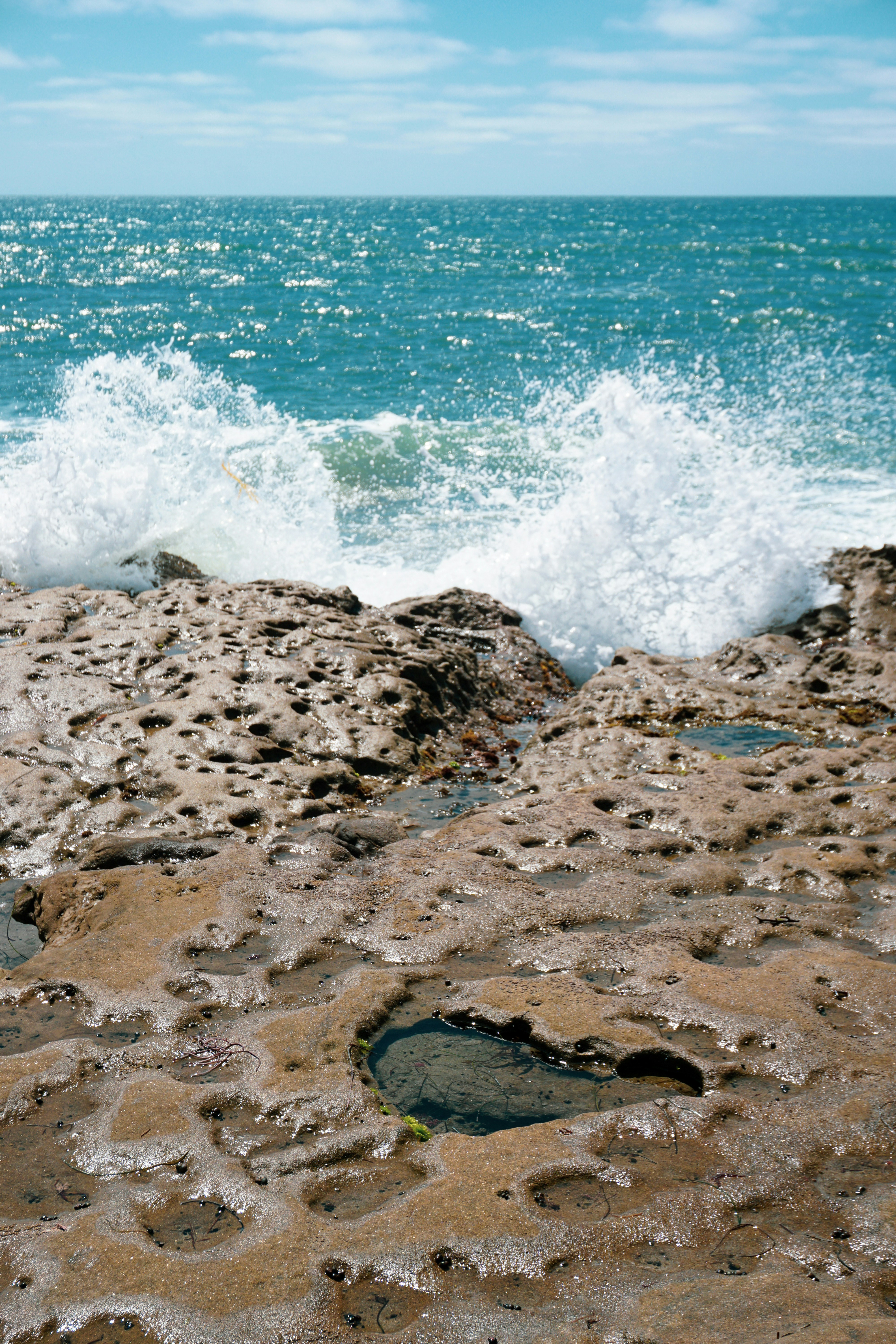 Rocky shoreline with tidal pools reflecting sunlight, as waves crash against the shore in a vibrant coastal scene.