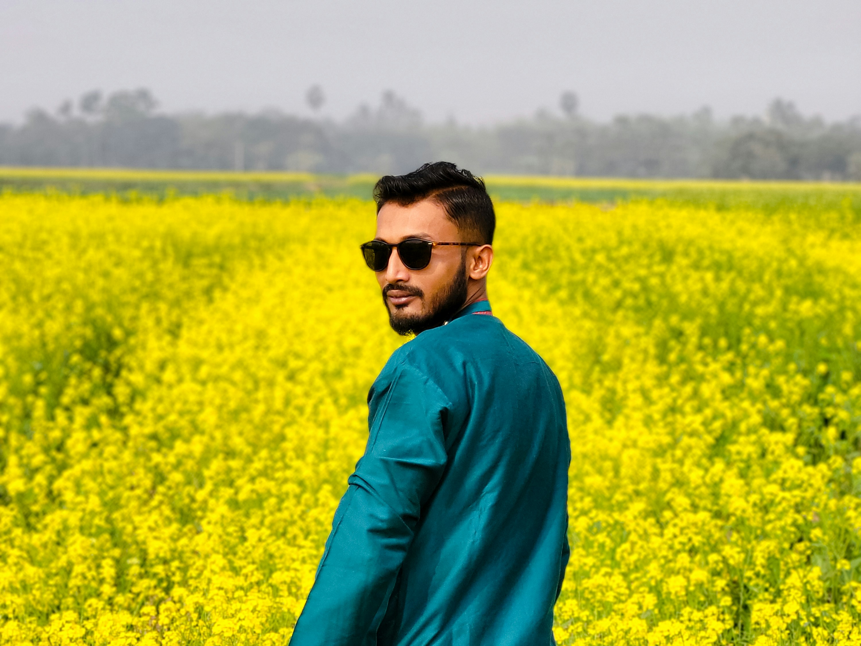 Man in sunglasses turning back while standing in a field of bright yellow flowers under a clear sky.