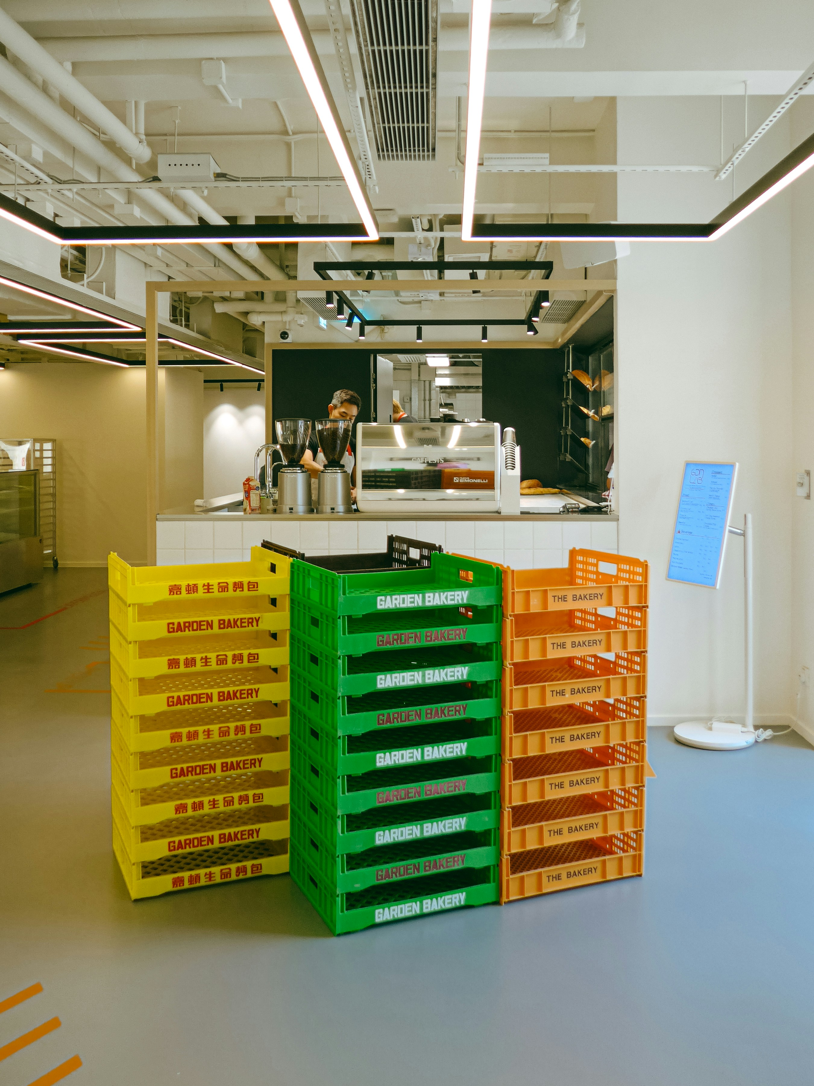 Bright industrial cafe with stacked colored crates forming a centerpiece; a coffee station sits behind, illuminated by geometric ceiling lights.