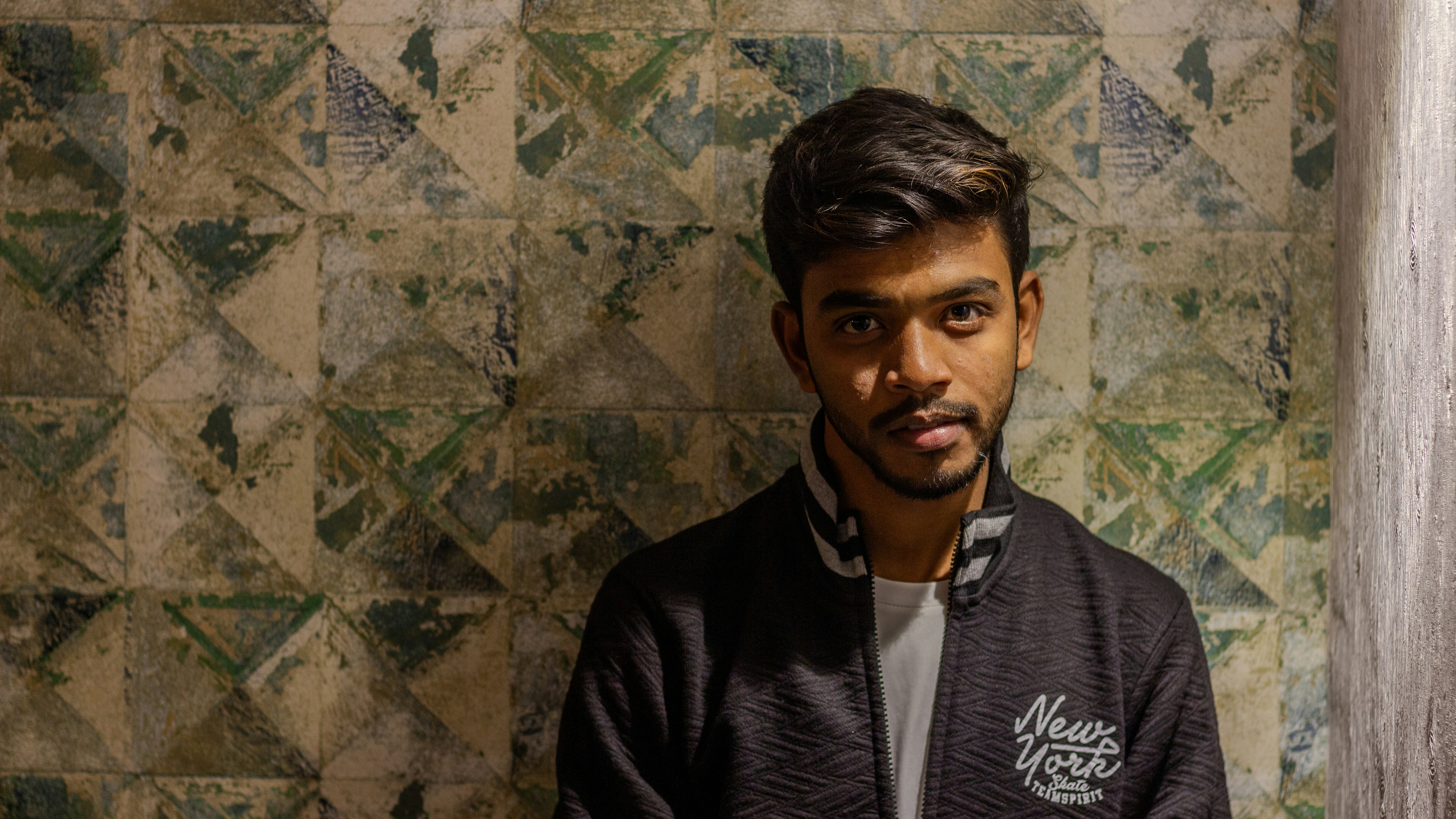 Man in a dark jacket stands against a textured wall with geometric patterns.