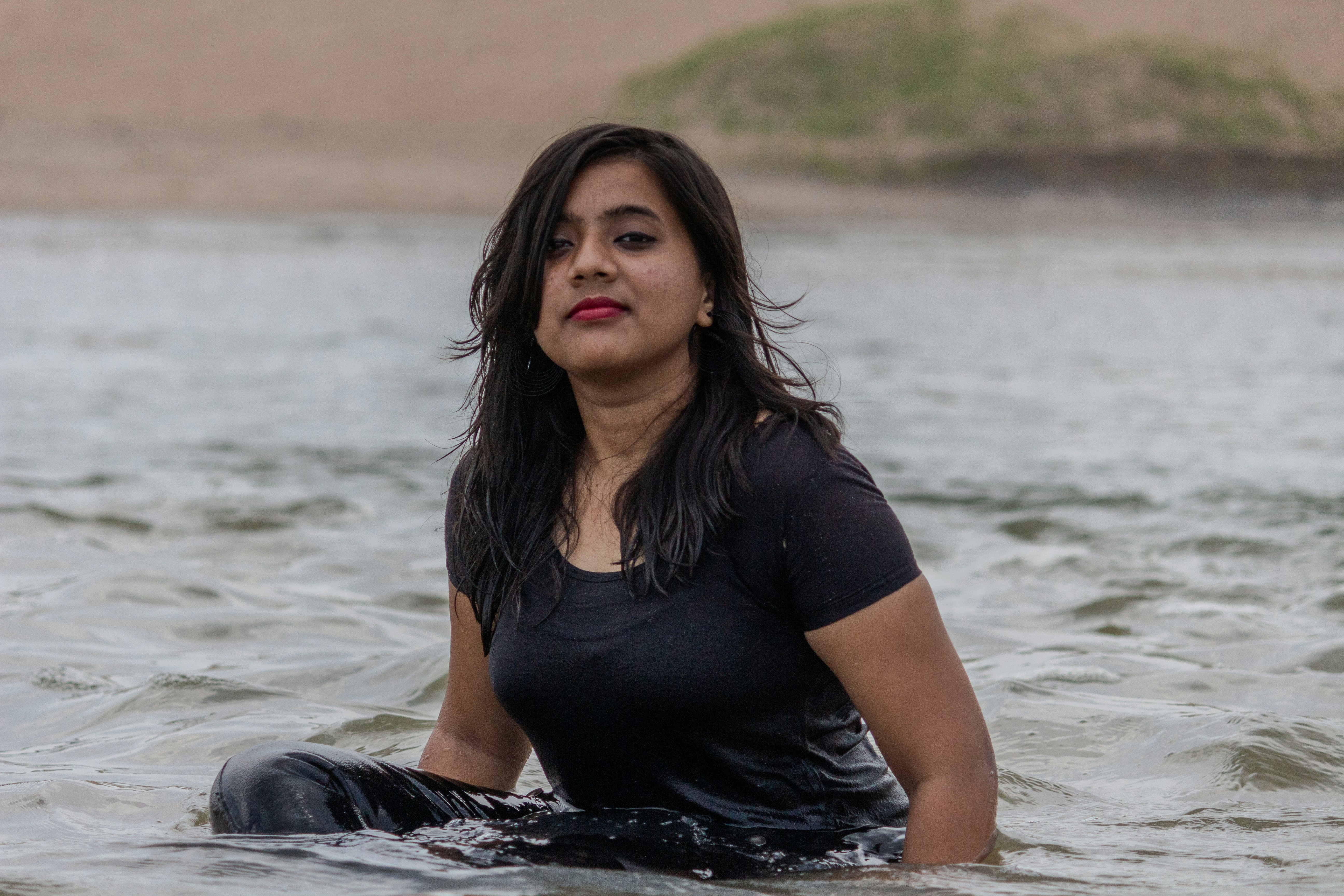 Woman seated in shallow water, surrounded by gentle waves and a sandy backdrop.