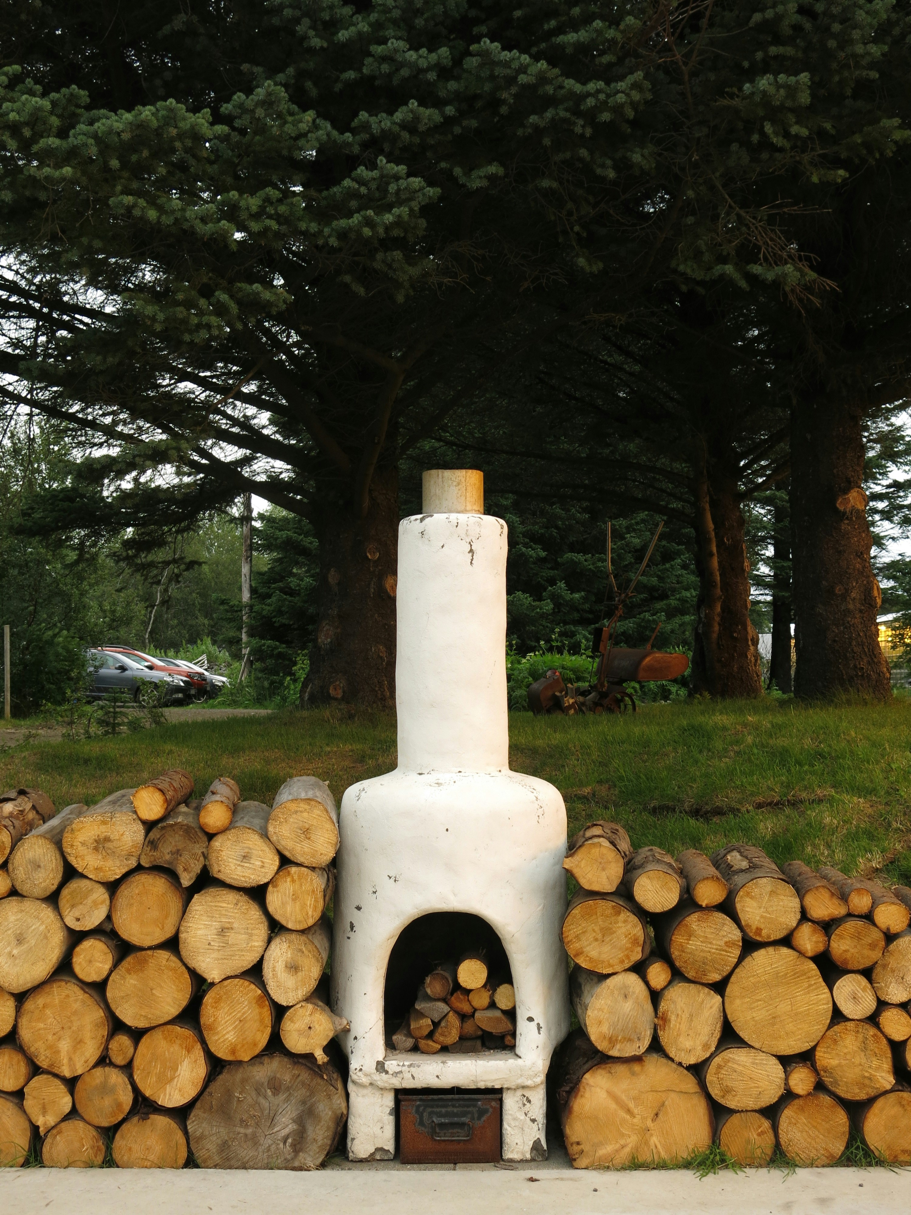 A white outdoor chimney stands proudly in front of a neatly stacked pile of logs, framed by lush greenery and towering trees.