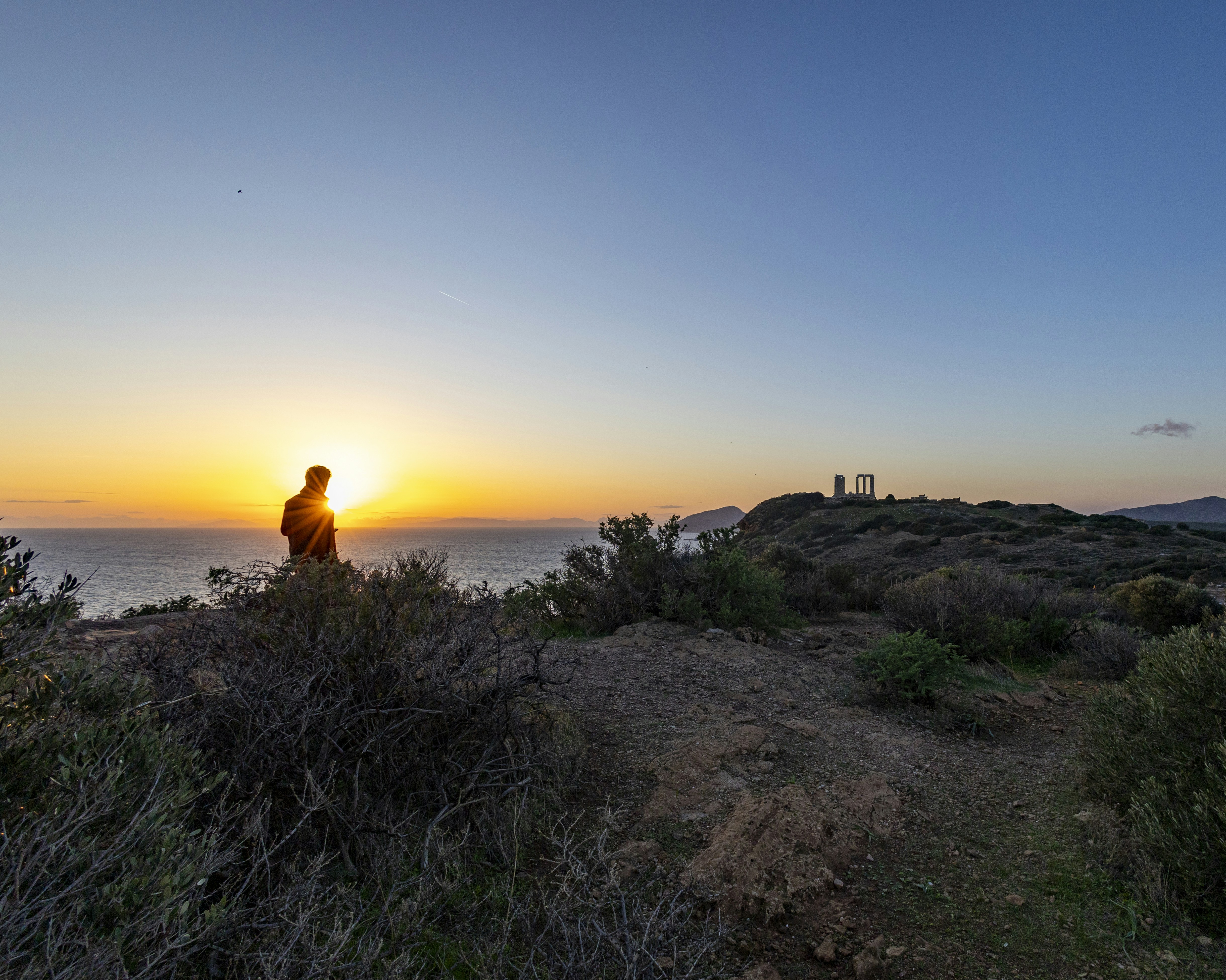 Person standing on a hillside at sunset with ancient ruins in the distance.
