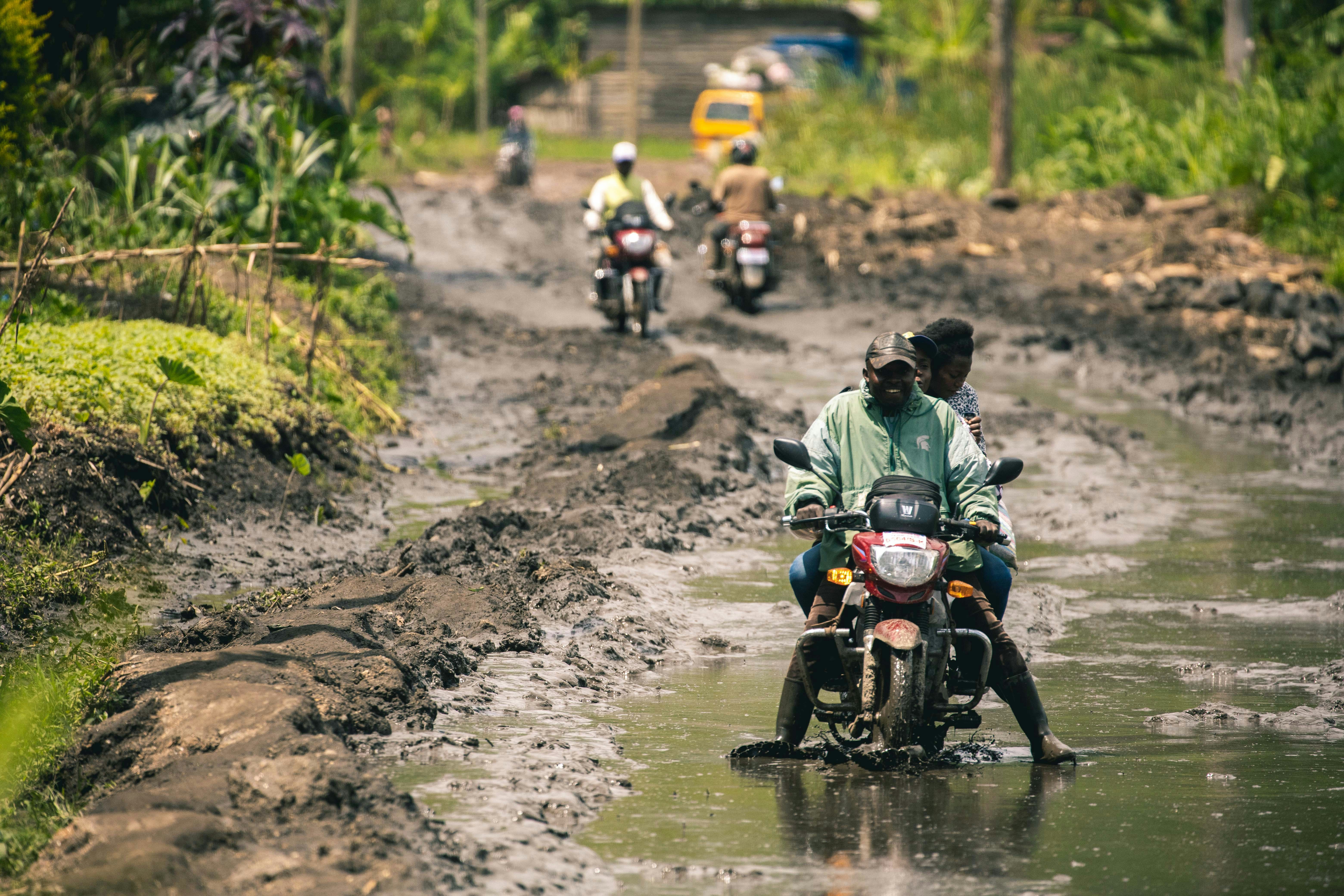 Motorcyclists maneuvering through a muddy road in a lush, tropical landscape, showcasing the challenges of rural transportation.