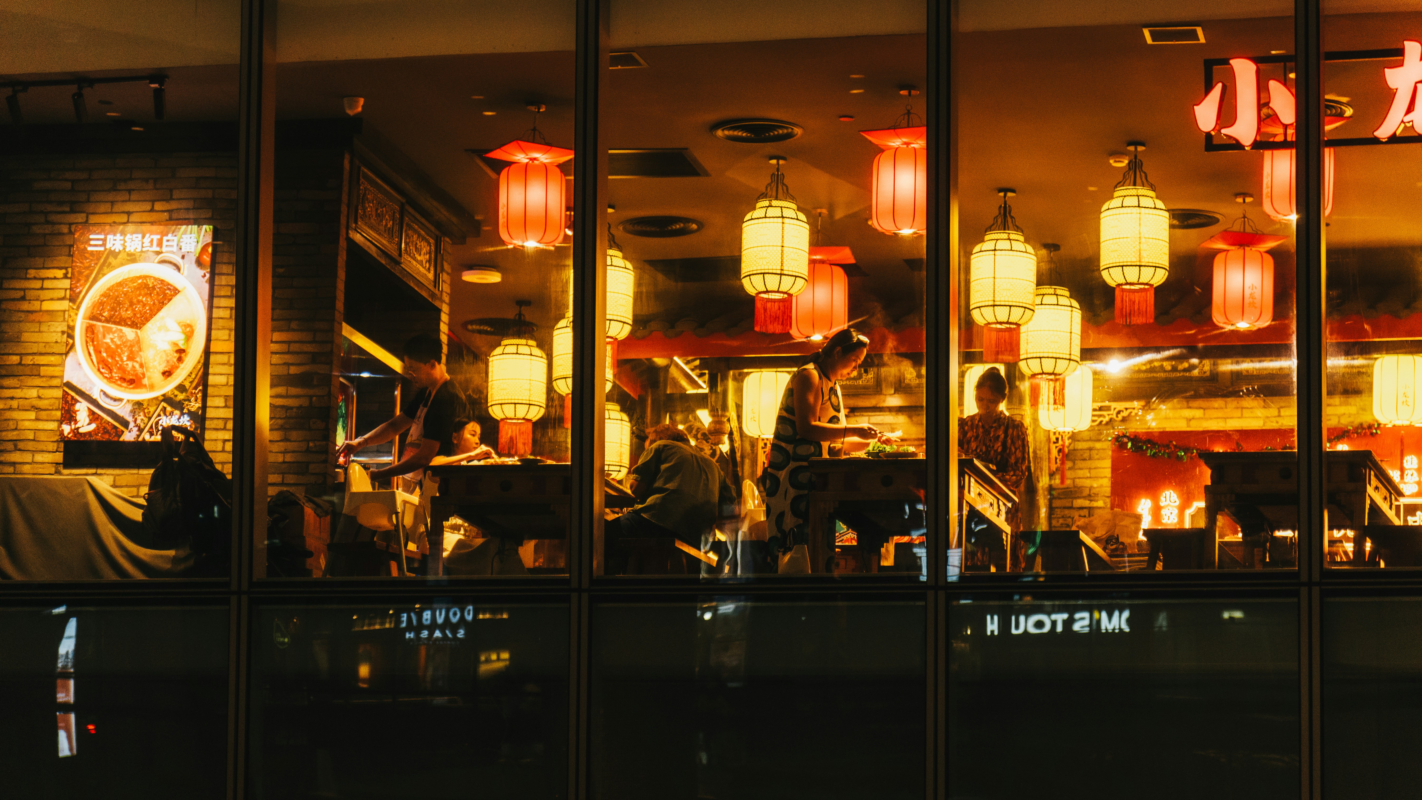 Evening dining scene through a glass window with red and yellow lanterns illuminating the interior.